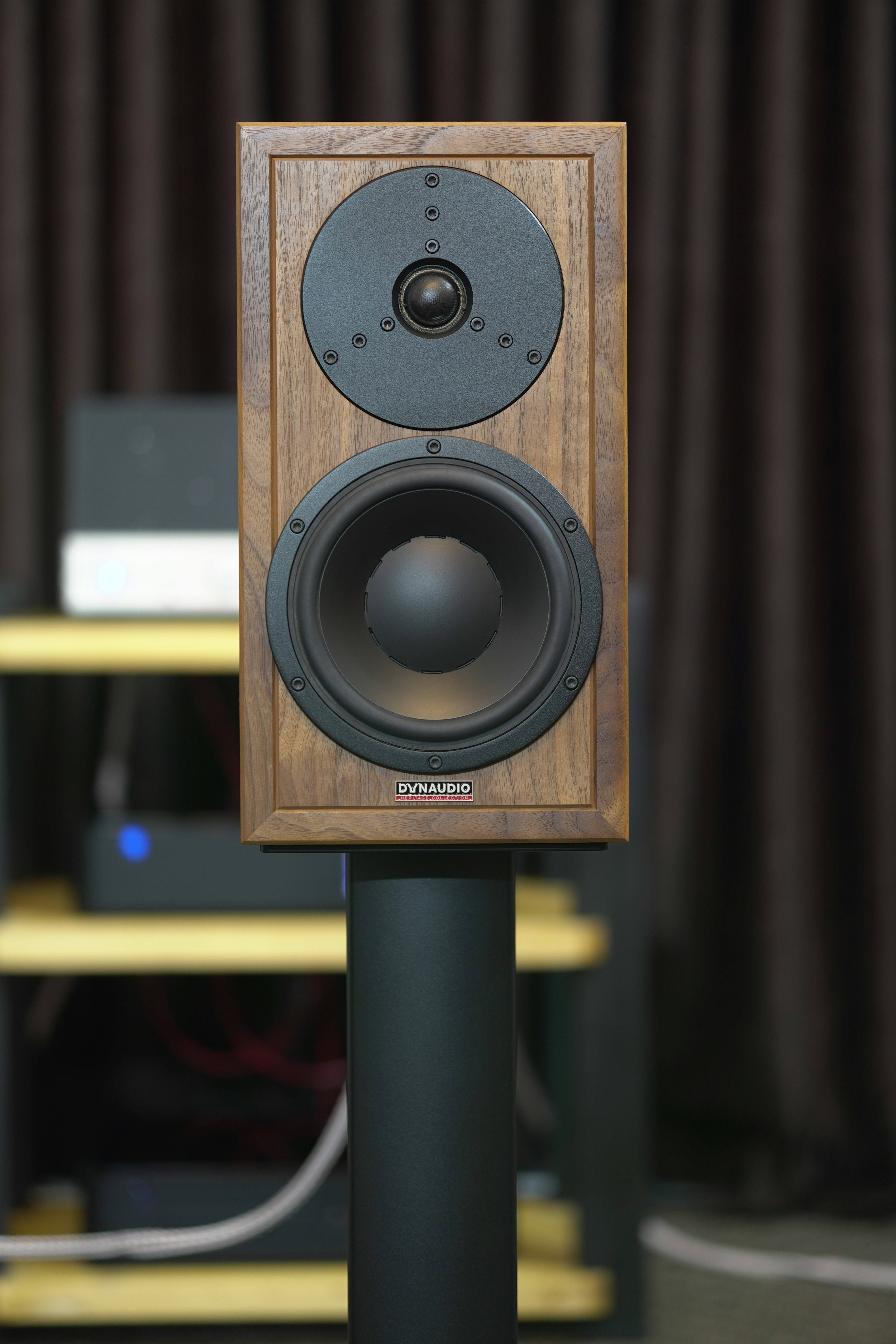 Dynaudio speaker on a stand, showcasing its elegant wood finish and dual drivers against a blurred background of audio equipment.