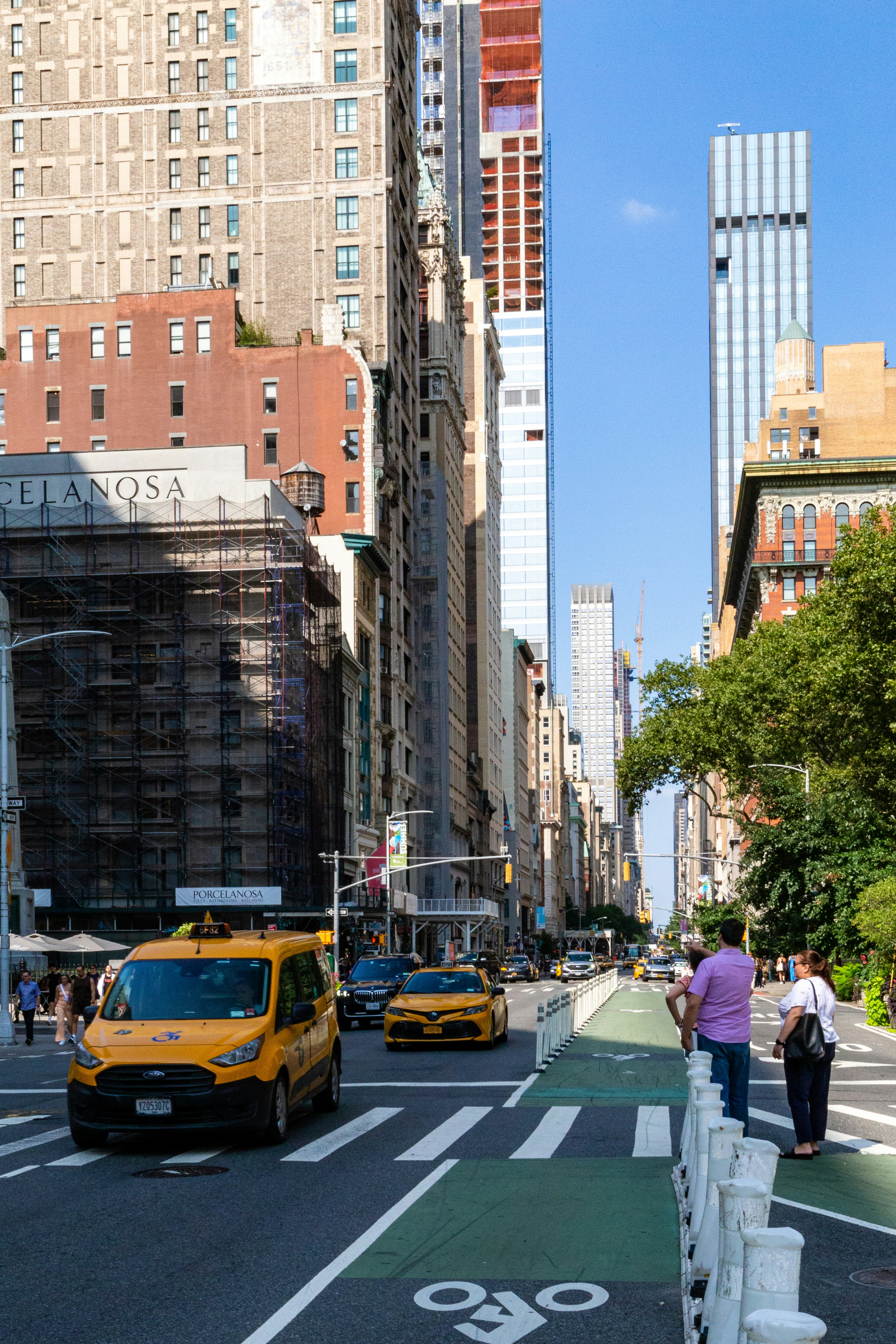 Busy city street showcasing yellow cabs navigating through traffic, with modern skyscrapers and lush greenery lining the road.