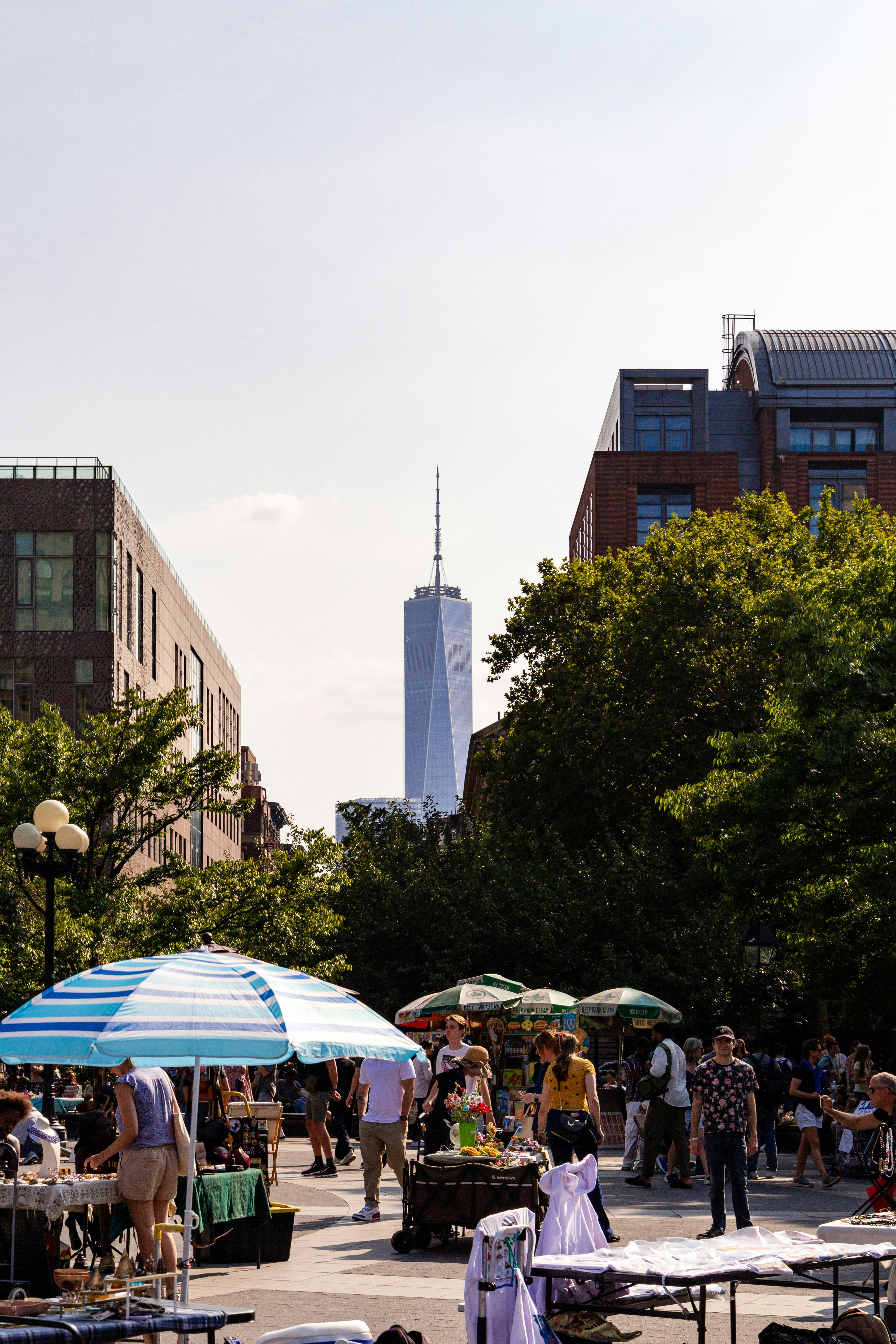 Vibrant street market bustling with activity under colorful umbrellas, framed by modern architecture and the One World Trade Center in the background.