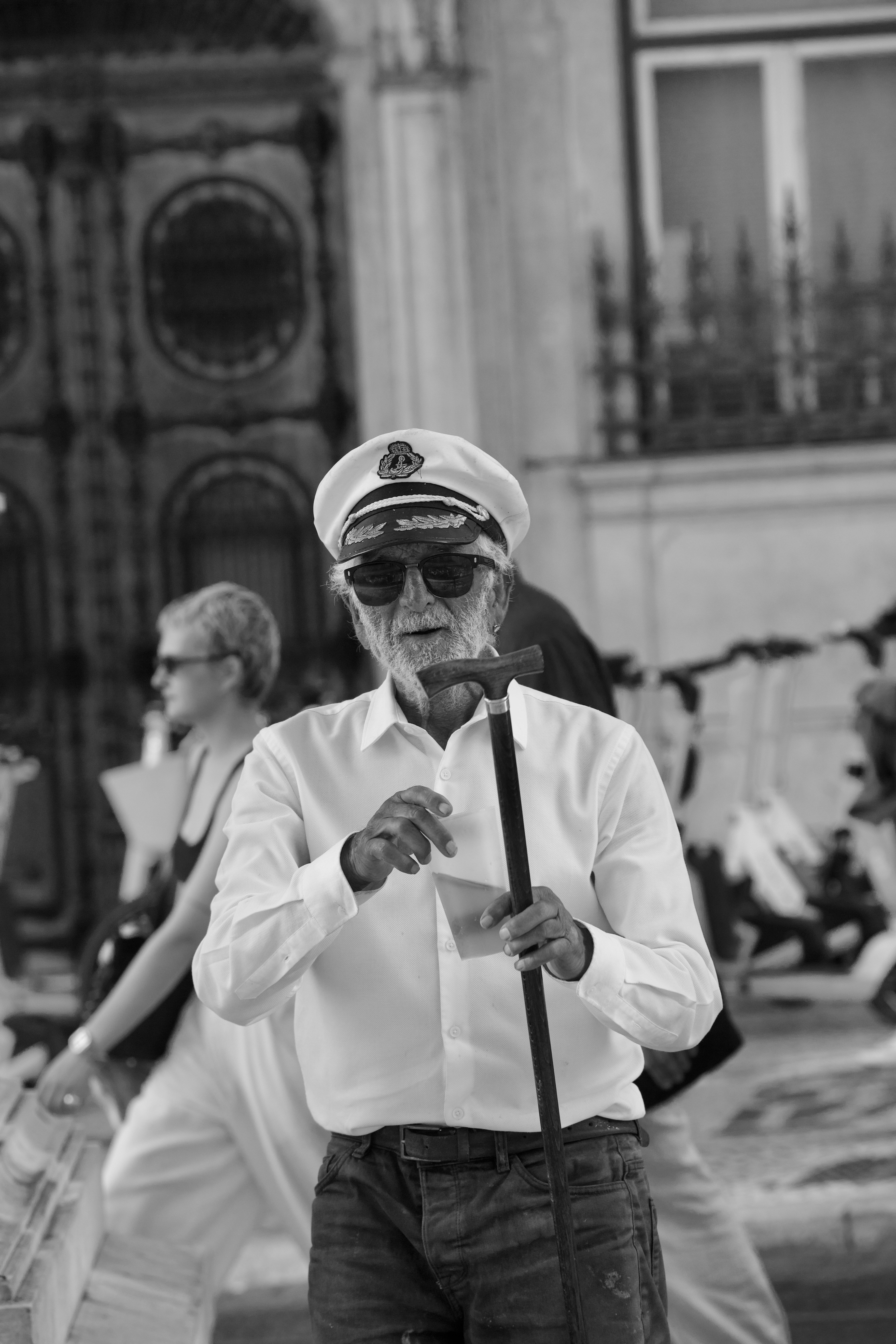Elderly man in a captain's hat and sunglasses, holding a cane, stands amidst a busy street scene. The background features blurred figures and historic architecture.