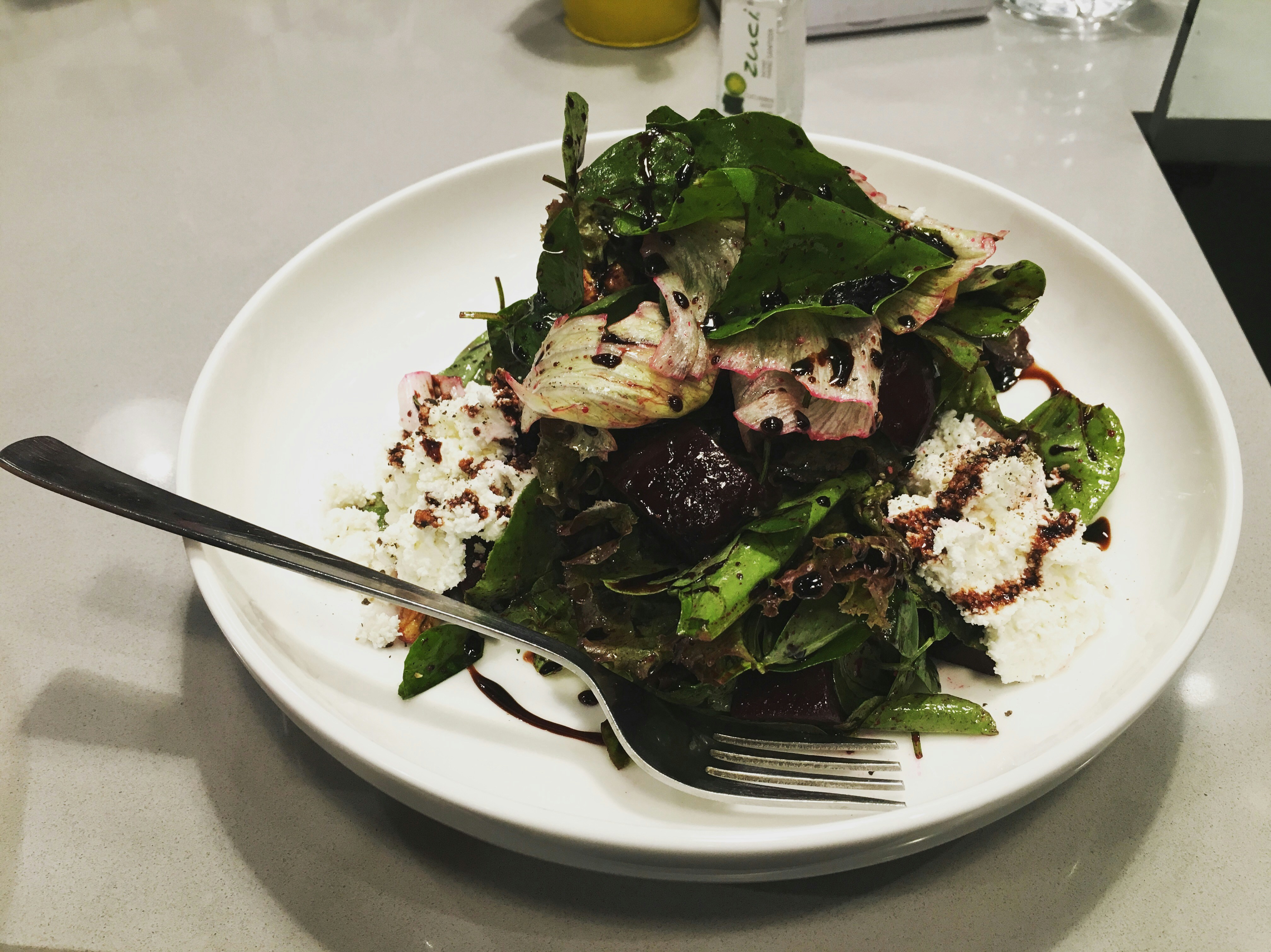 Vibrant salad featuring mixed greens, beets, and dollops of creamy cheese, artfully arranged in a white bowl. A fork rests beside it, inviting a taste.