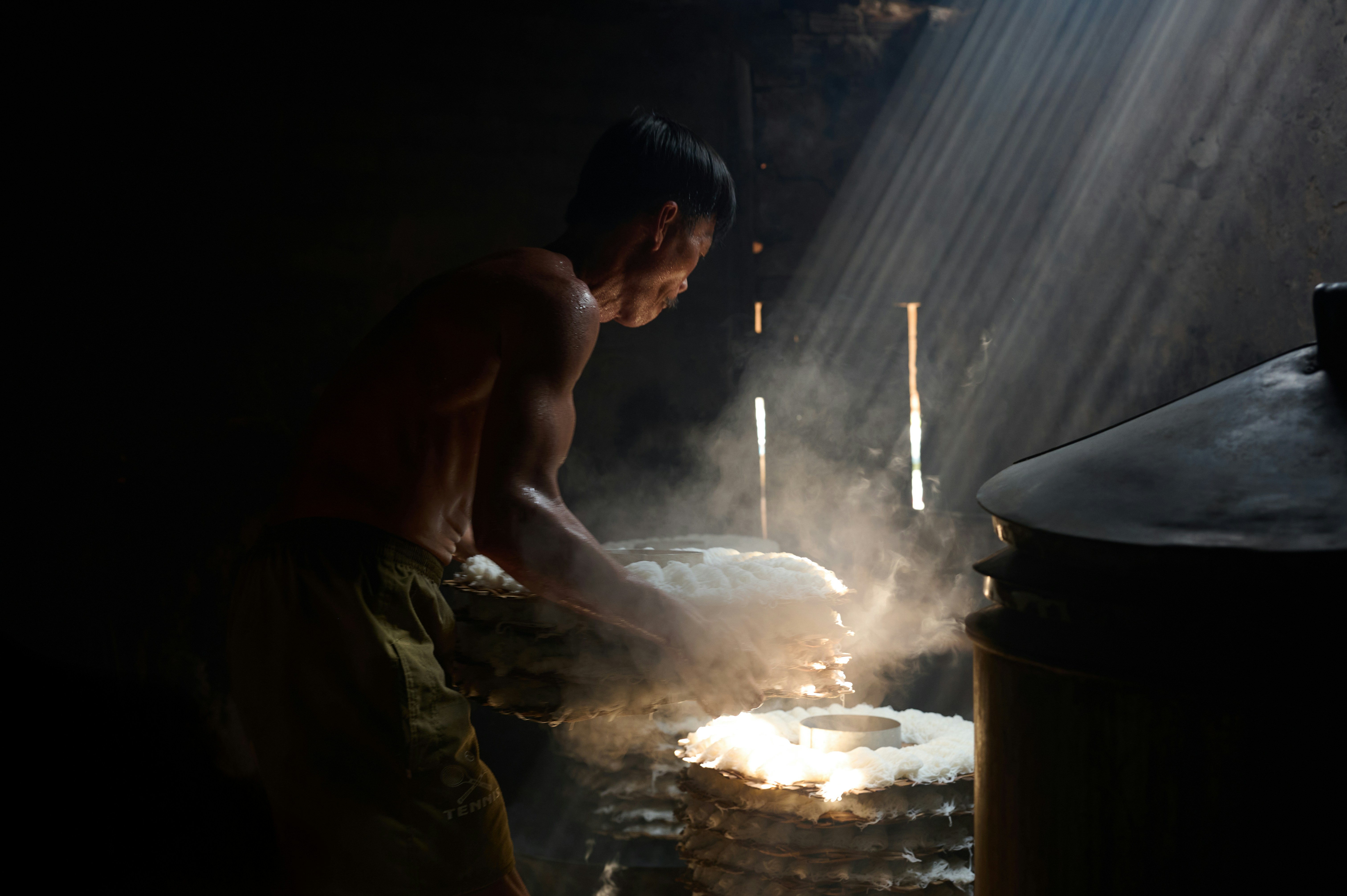 A worker carefully handles stacks of salt in a dimly lit environment, illuminated by shafts of light breaking through the darkness.