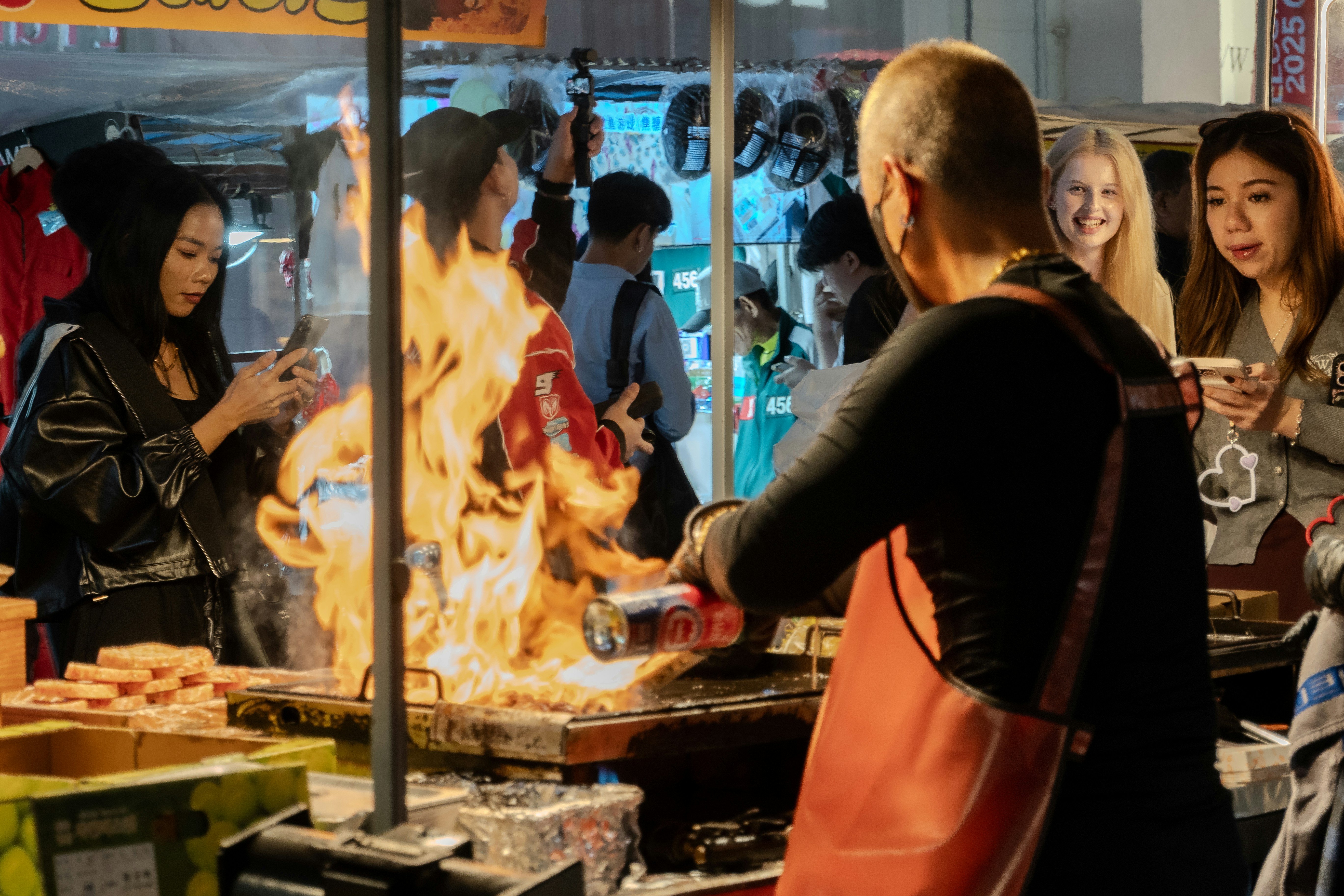 A vendor skillfully prepares food over an open flame, surrounded by intrigued onlookers at a bustling night market.