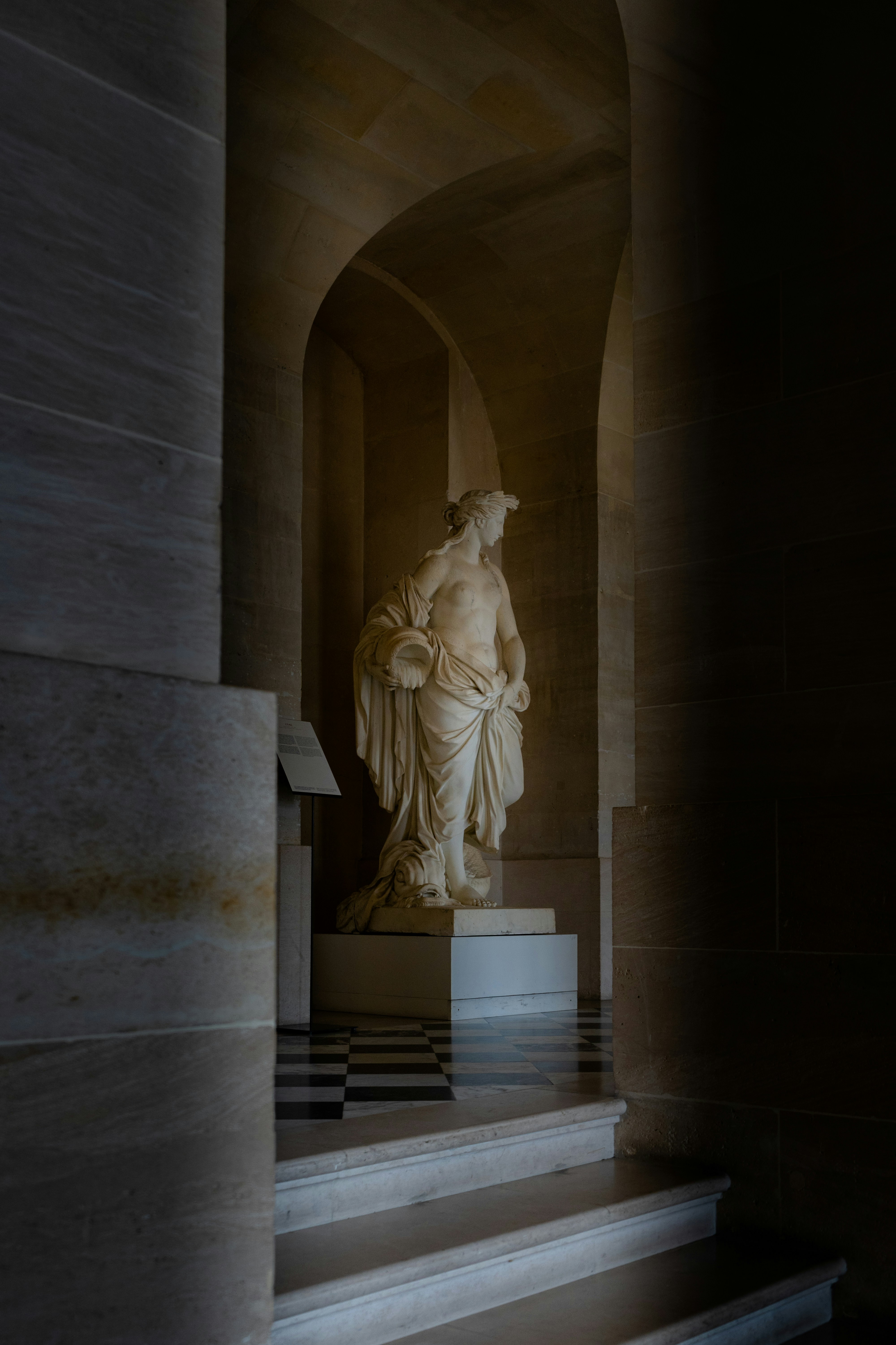 White marble statue of a figure draped in flowing robes, positioned in a softly lit alcove with a checkered floor.