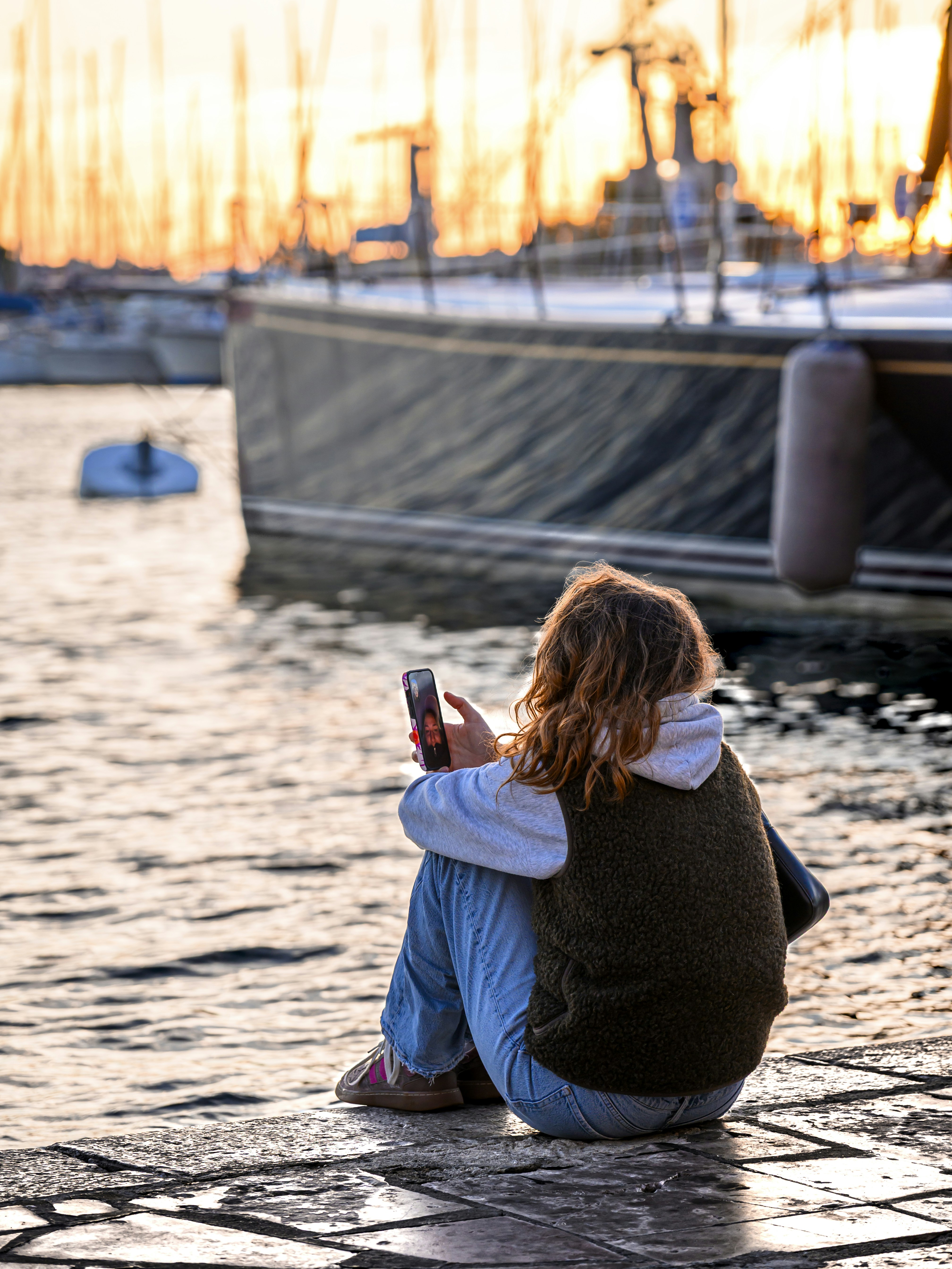 Woman with phone by water with boats at sunset