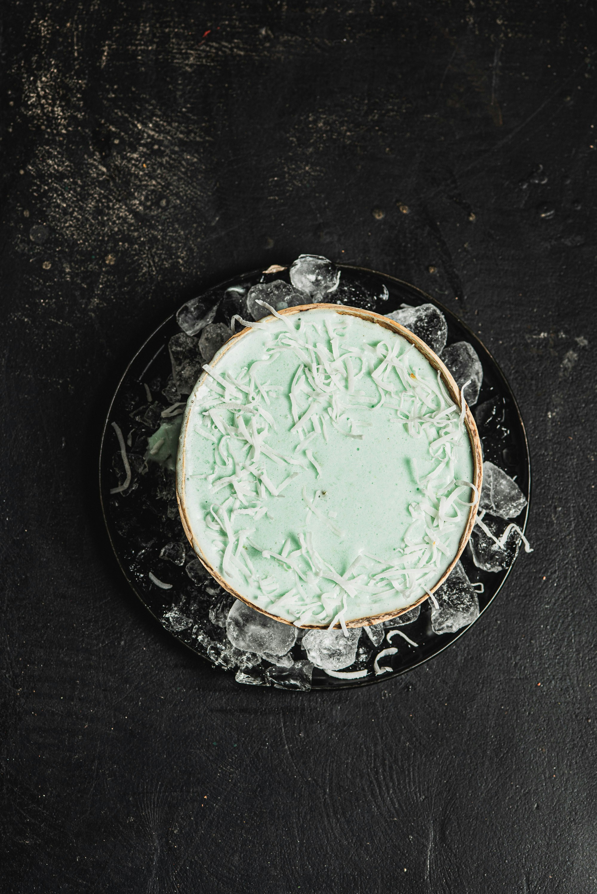 A mint-flavored dessert topped with shredded coconut, resting on a bed of ice cubes against a dark backdrop.
