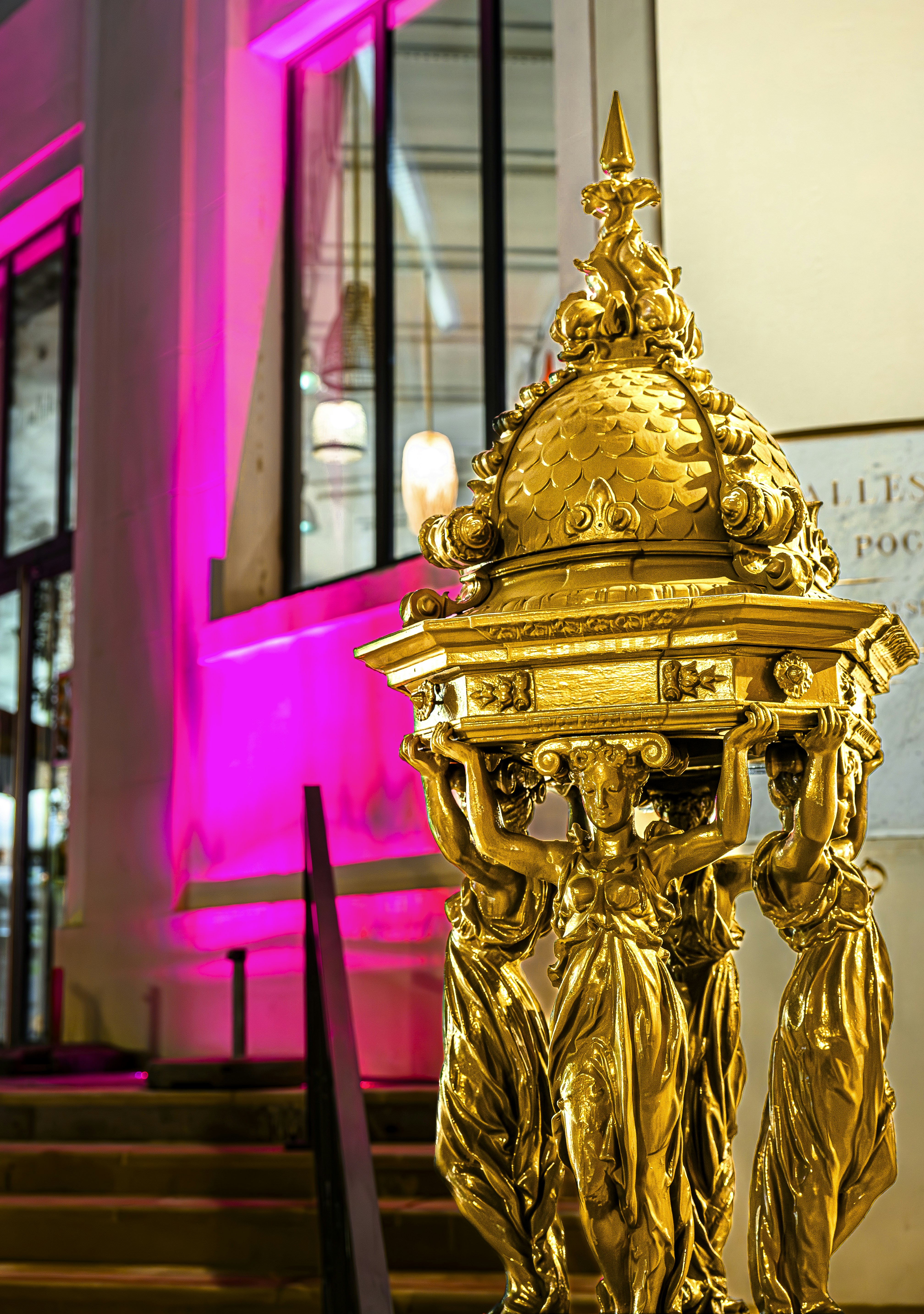 Ornate golden sculpture supported by four figures, illuminated by soft pink lights in a contemporary setting.