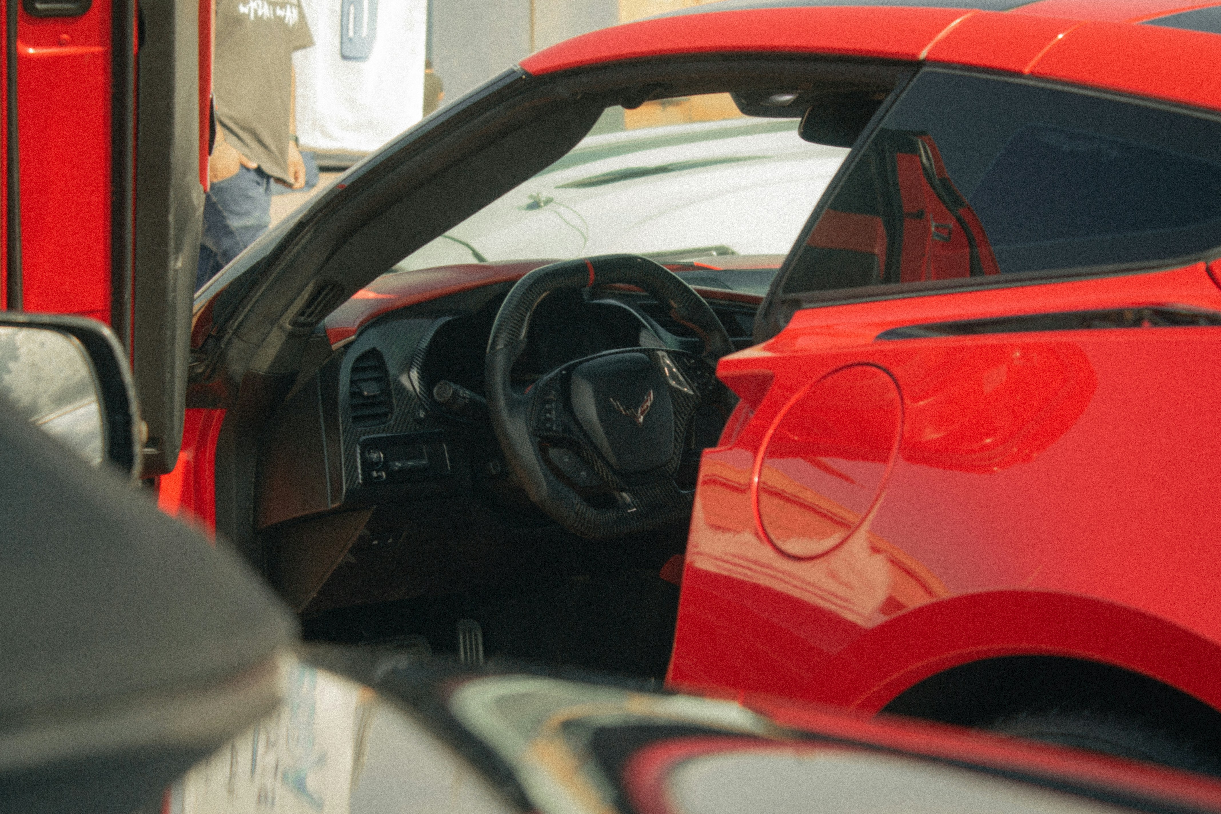 Interior view of a red Corvette showcasing the steering wheel and dashboard, with reflections hinting at a bustling environment outside.