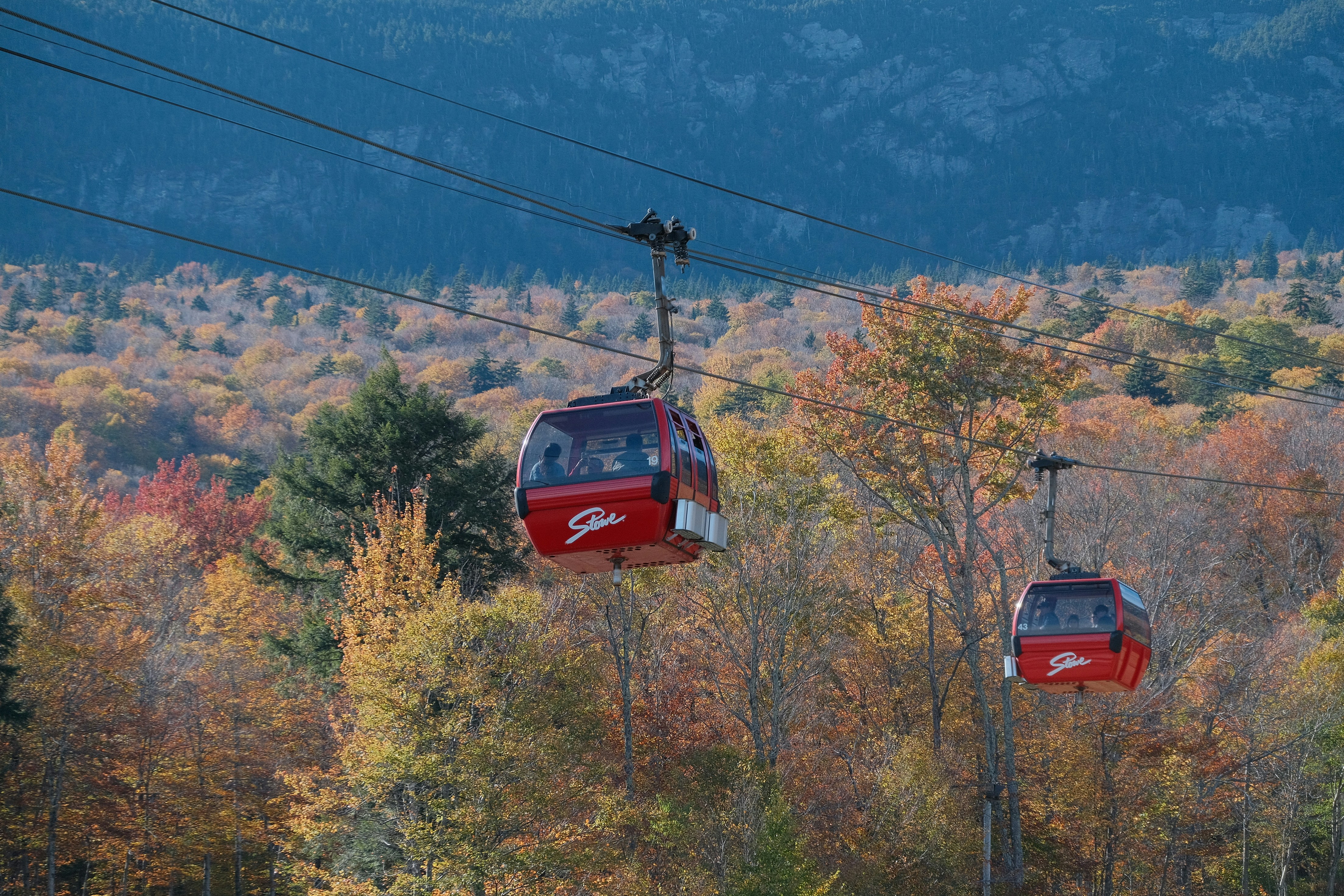 Two red gondolas glide above a vibrant autumn landscape, showcasing a tapestry of orange and yellow foliage against a mountainous backdrop.