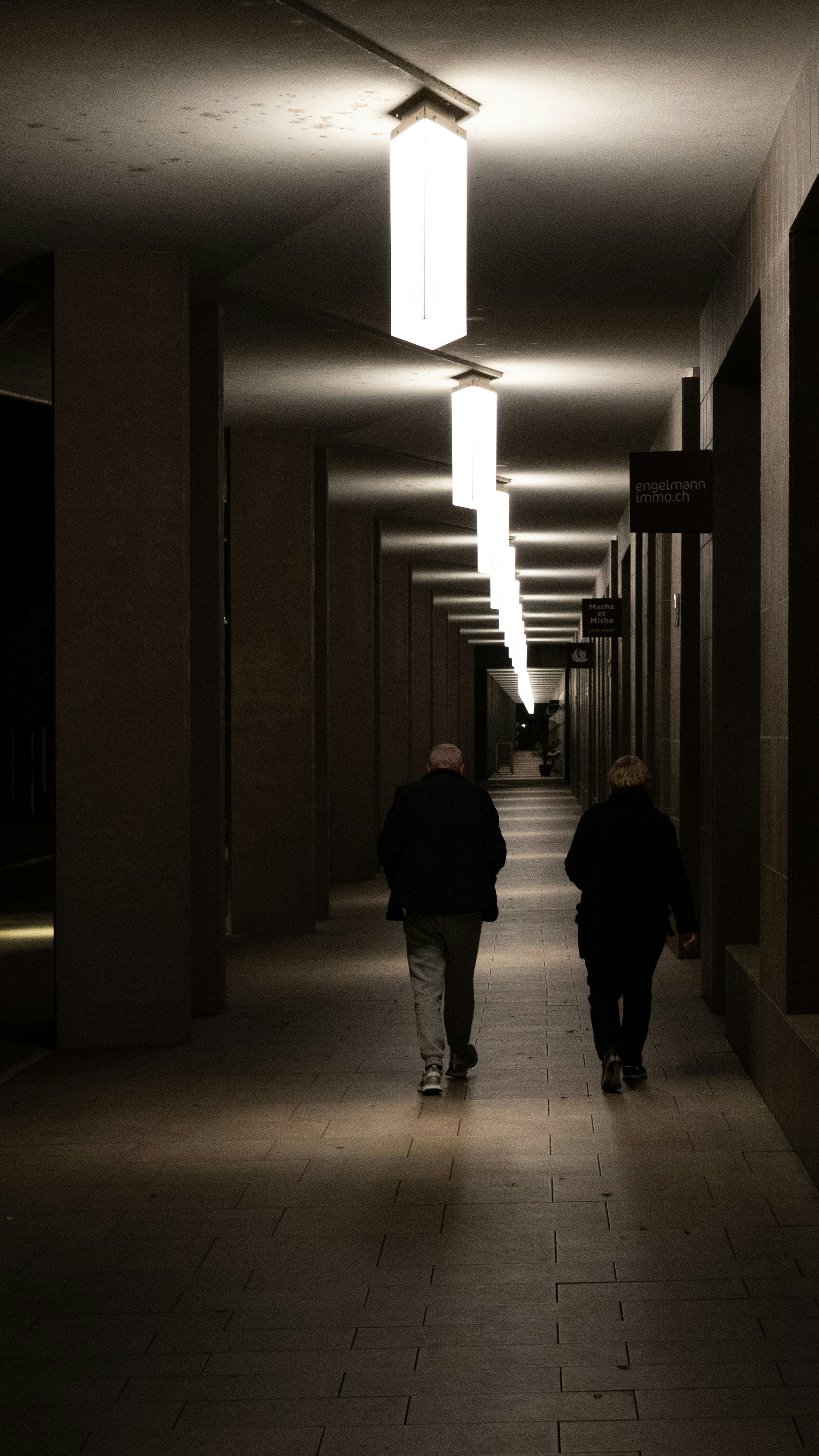 Two people walk down a dimly lit colonnade at night.