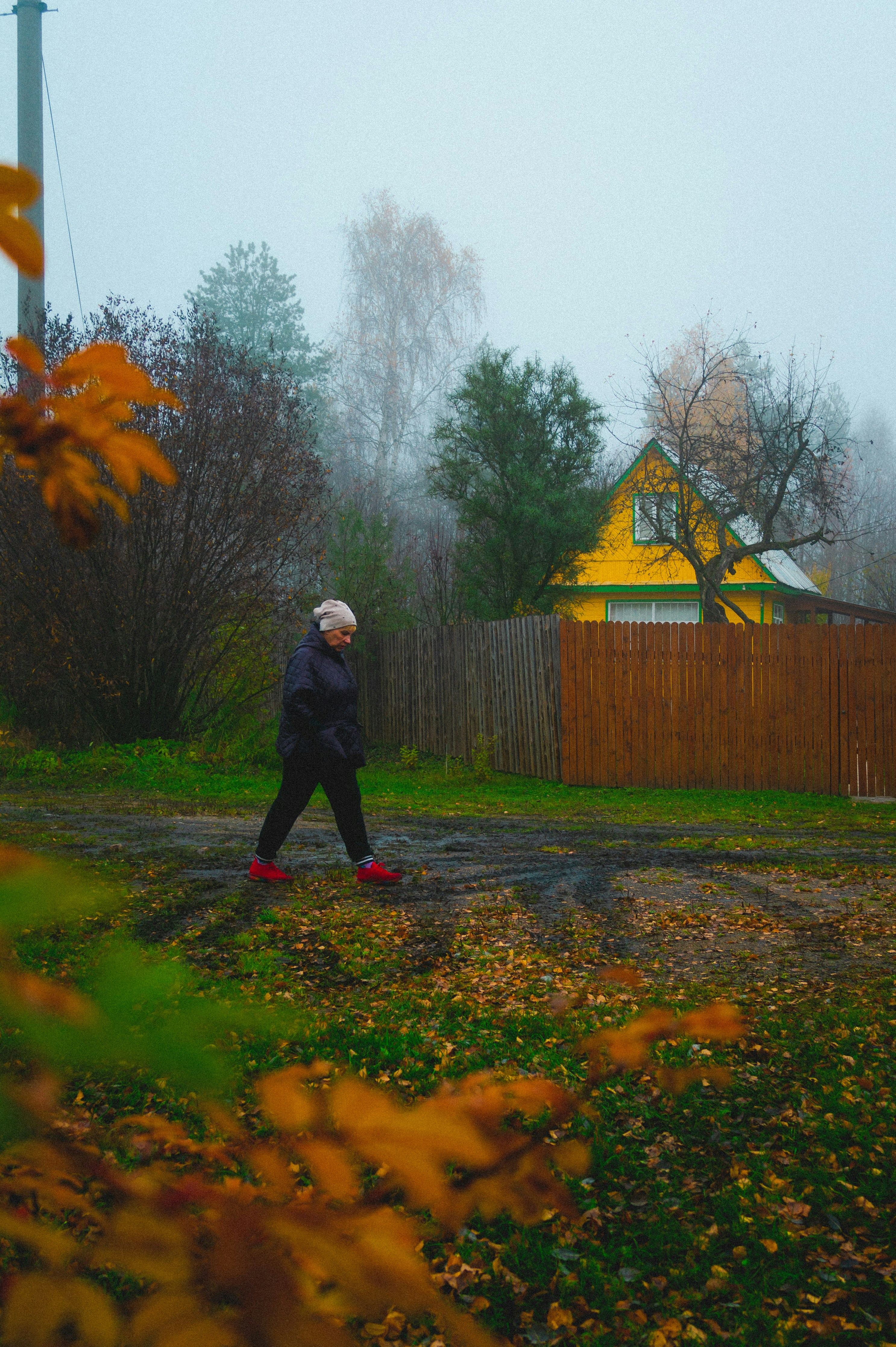 Elderly woman walking along a misty path, surrounded by autumn foliage and a vibrant yellow house in the background.