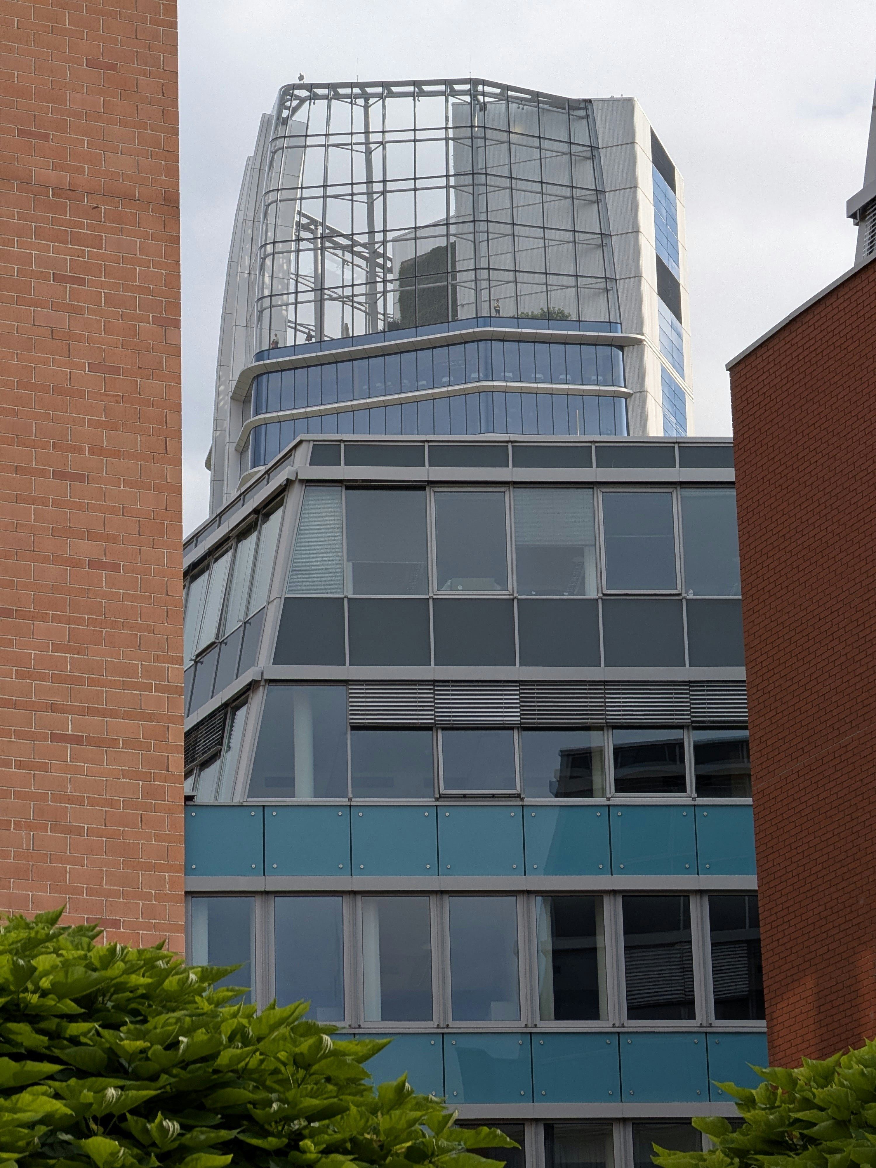 Modern glass building reflecting the sky, framed by brick structures and greenery.