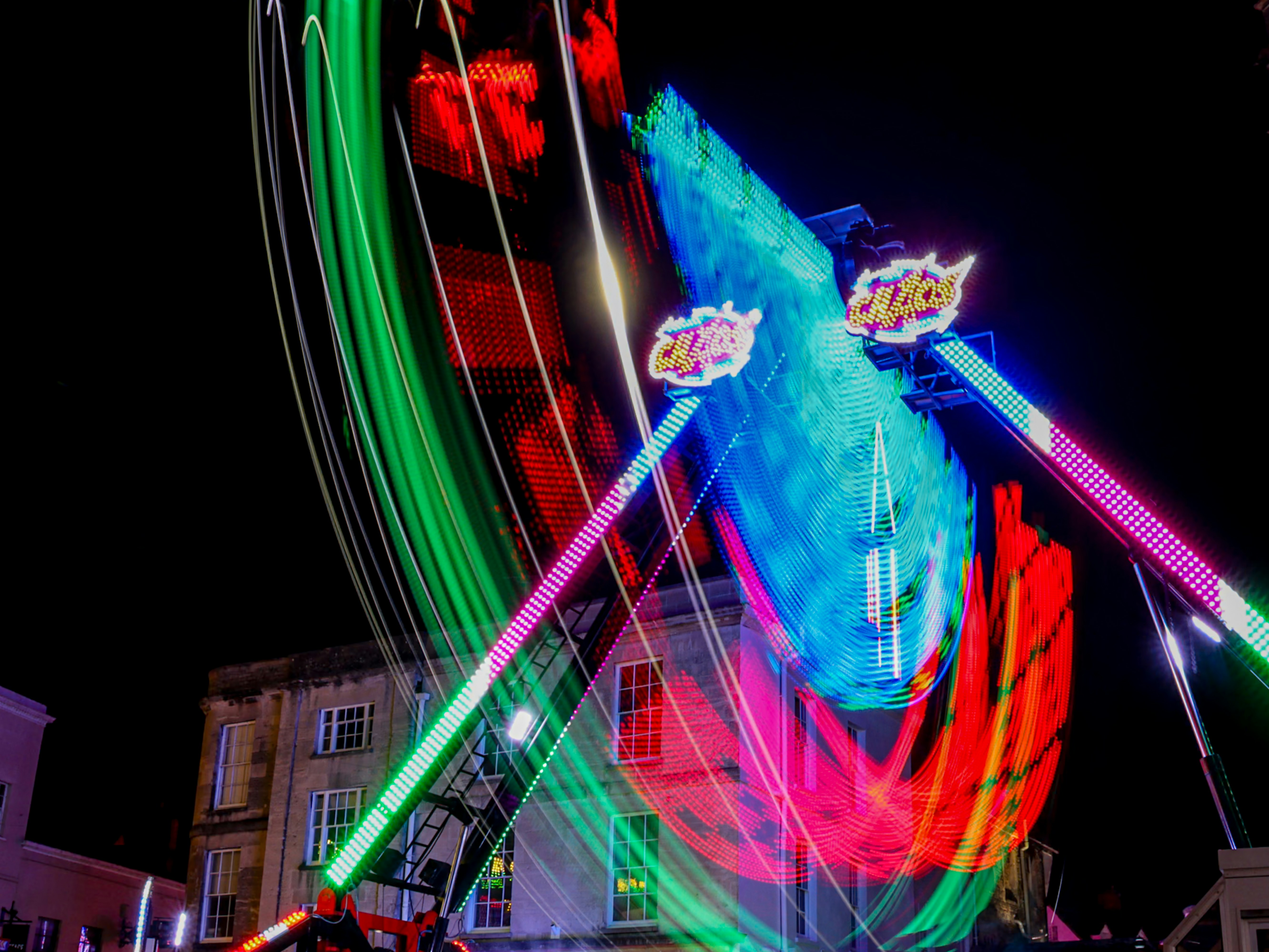 Dynamic light display from a carnival ride illuminating the night sky, showcasing a blend of colors and movement. The backdrop features historical architecture.