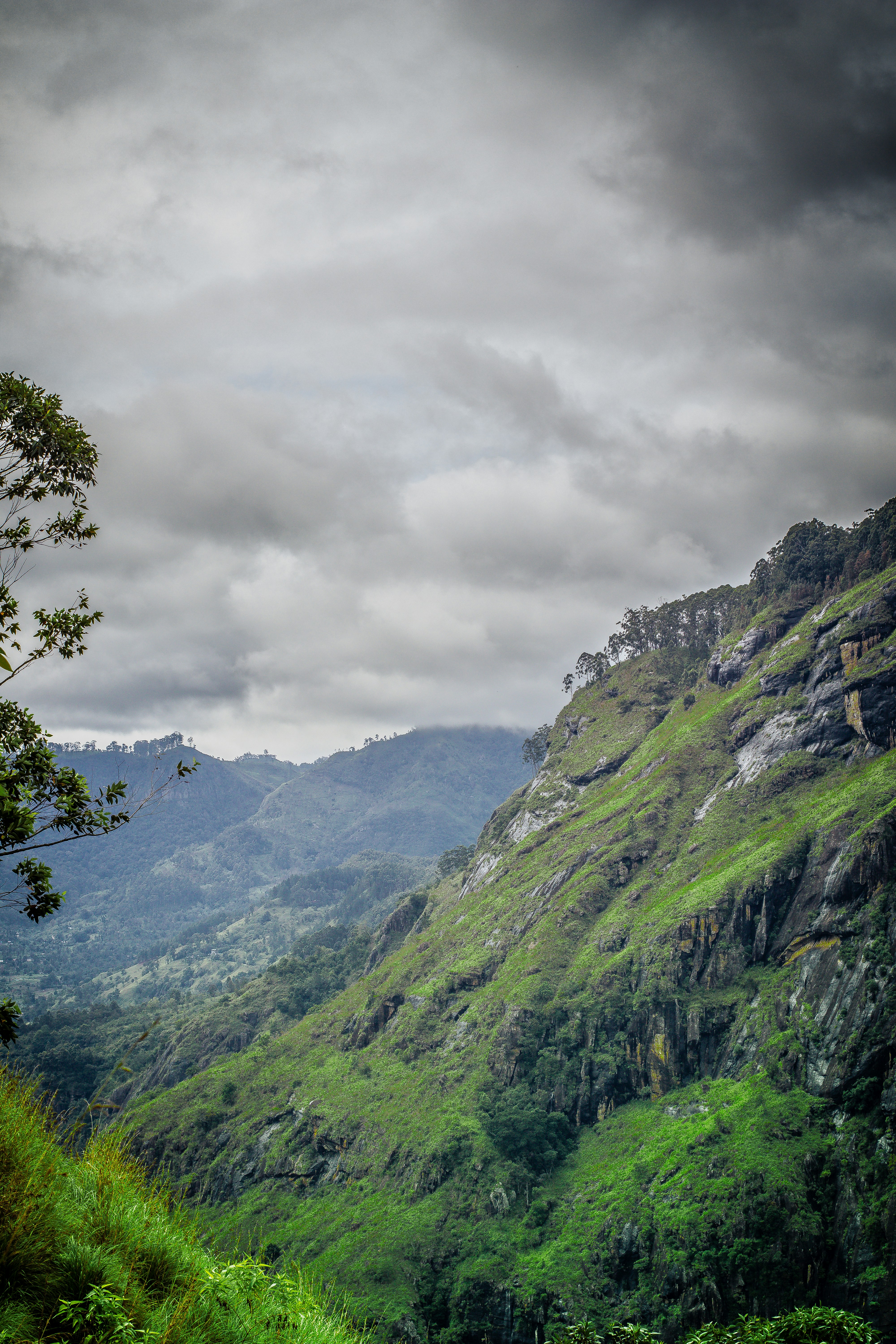 Lush green mountains rise dramatically beneath a moody sky, showcasing the rugged beauty of the landscape. The scene captures the interplay of light and shadow across the terrain.