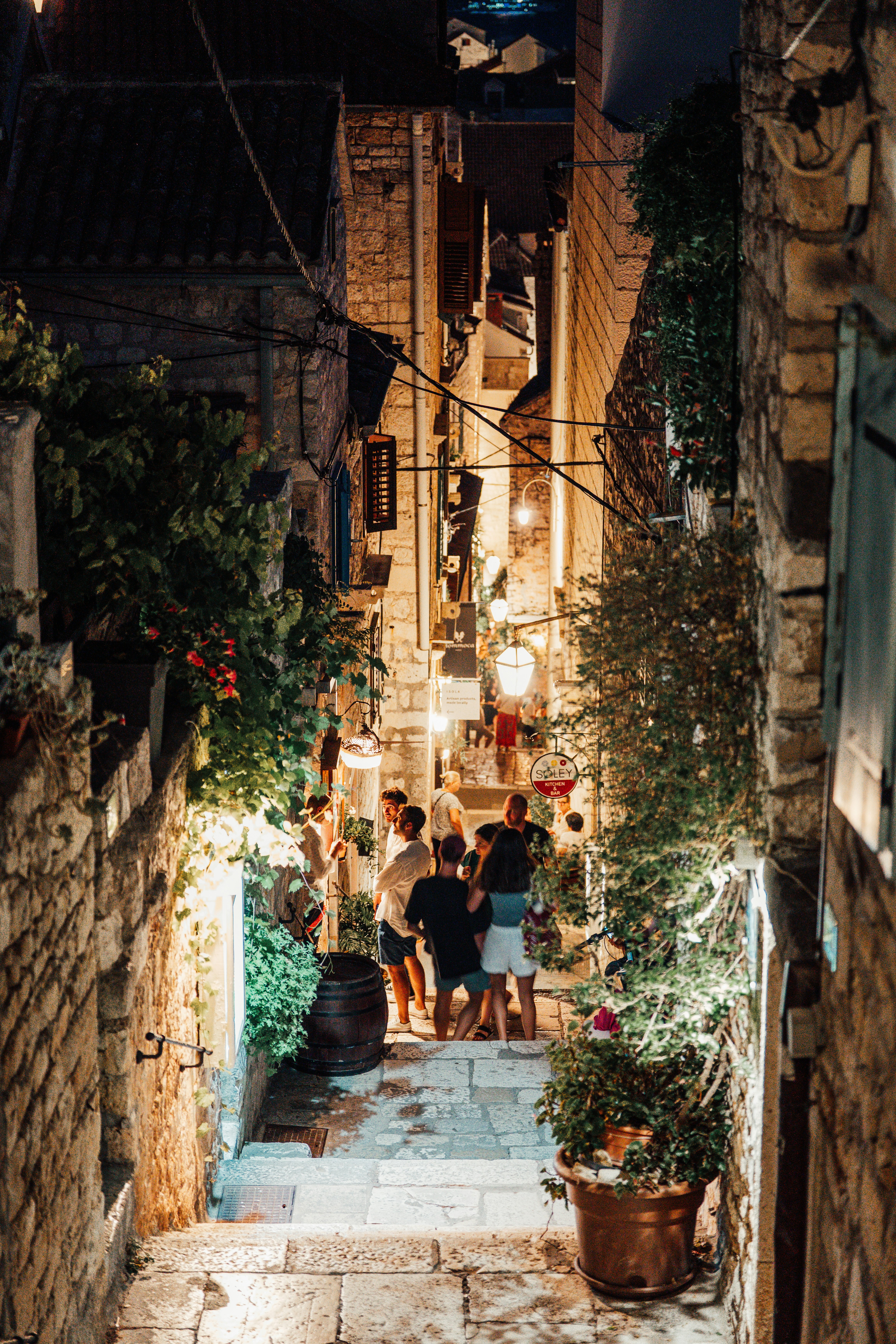 Charming narrow alley illuminated by warm lights, bustling with people enjoying the evening ambiance. Lush greenery and stone walls frame the scene.