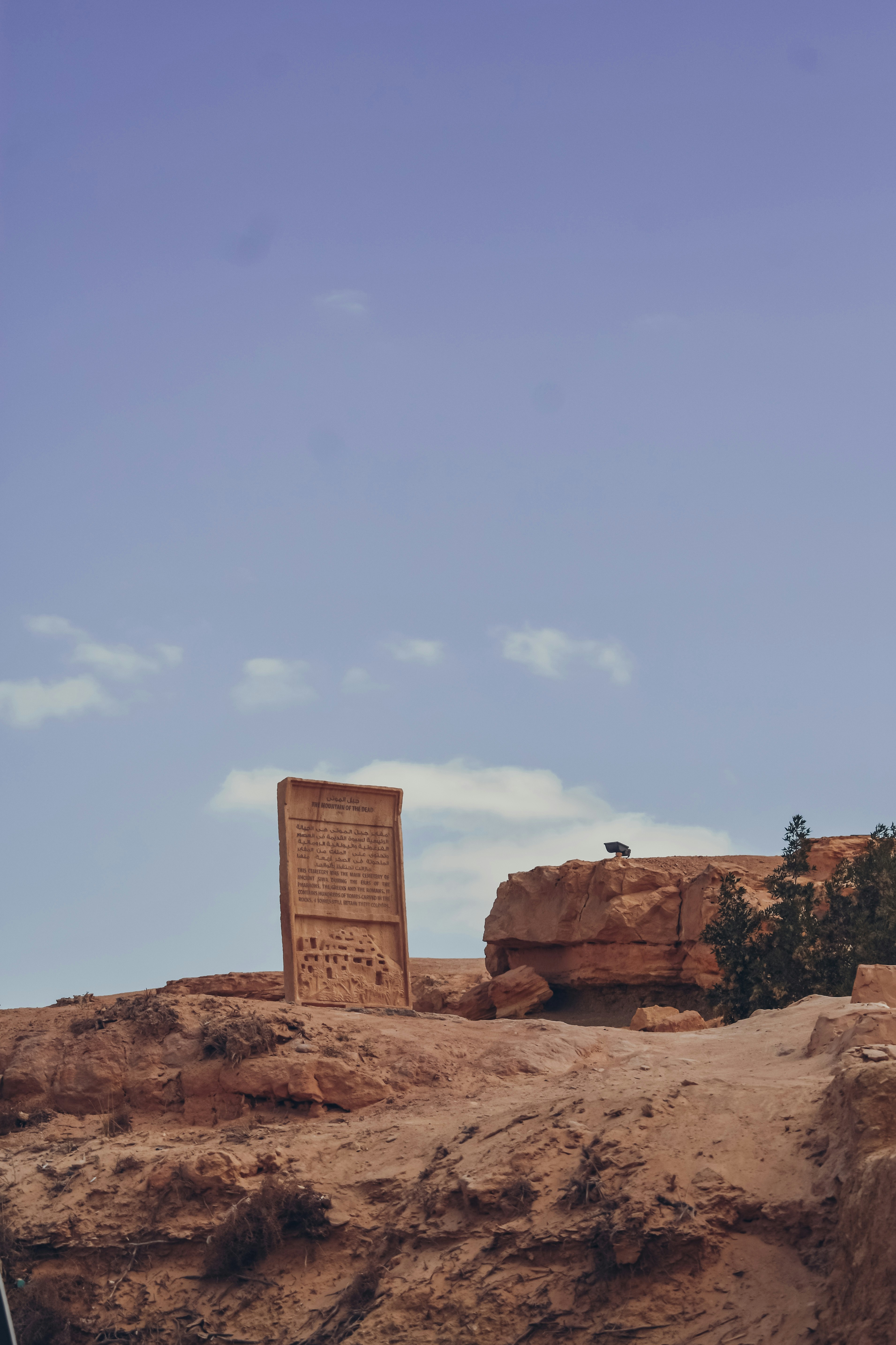 Weathered stone tablet stands amidst rugged terrain, surrounded by sparse vegetation under a clear blue sky.