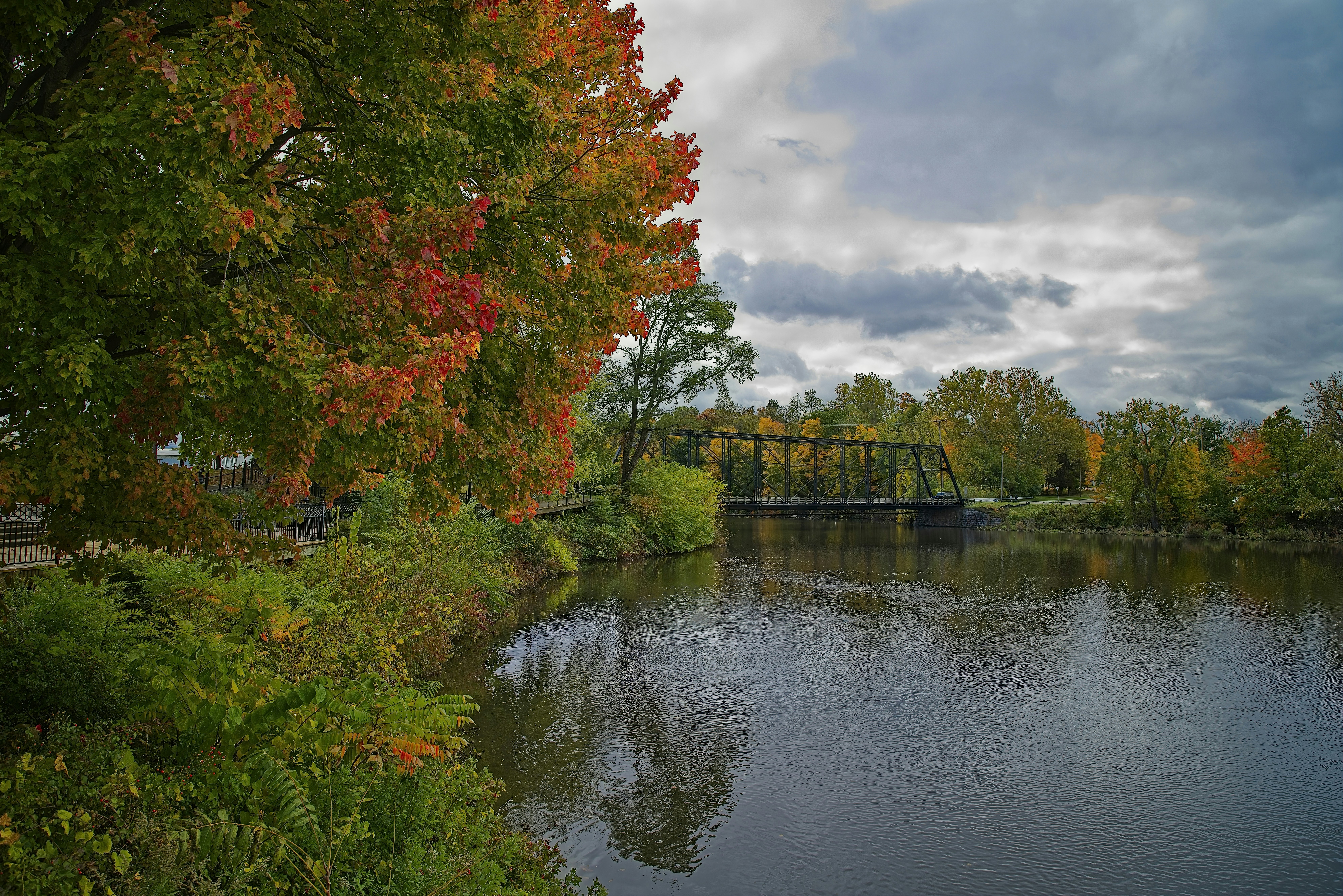 Vibrant autumn foliage frames a tranquil river scene, with a vintage bridge spanning the water under a moody sky.
