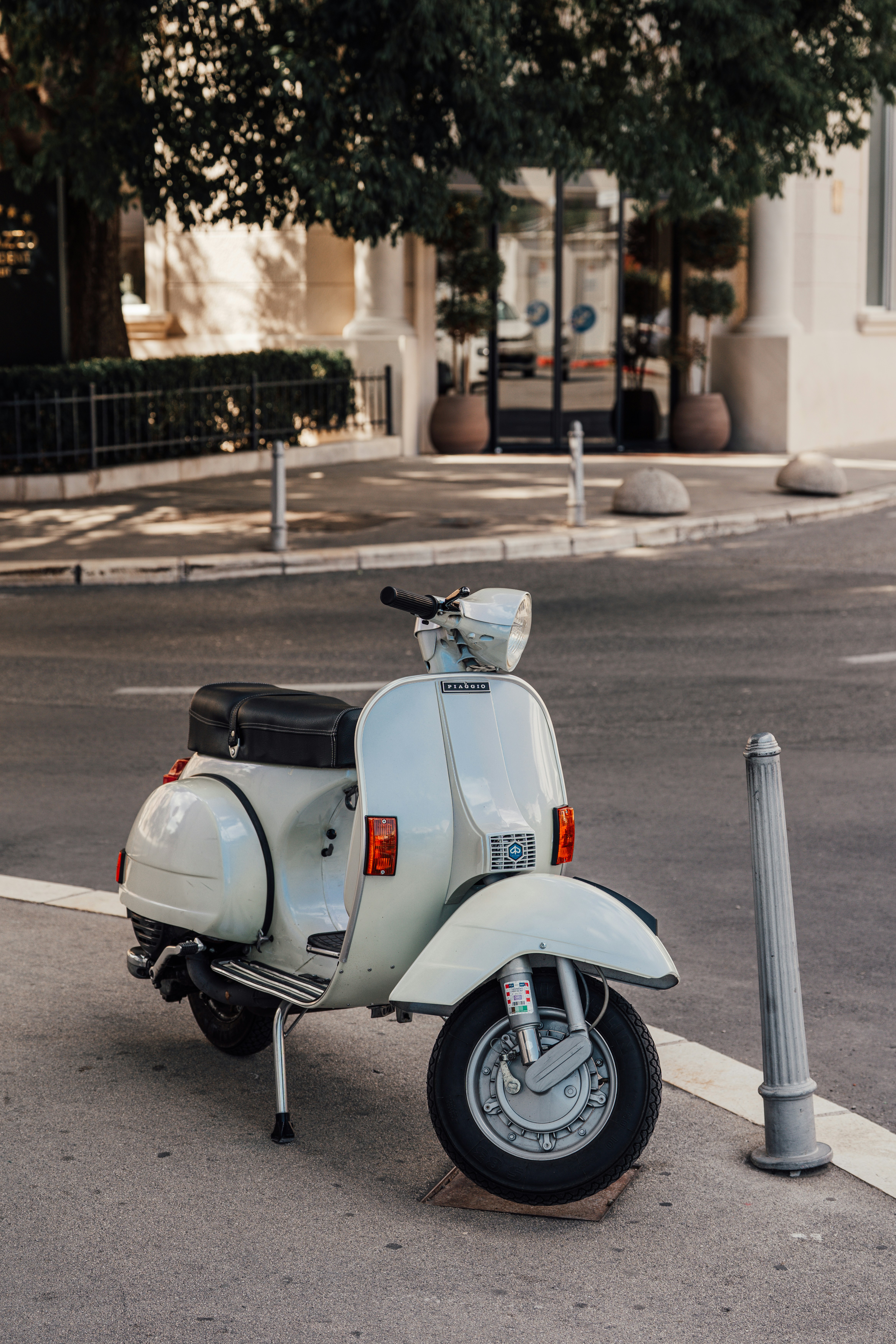 Classic light blue scooter parked on a city street, surrounded by urban elements and greenery.