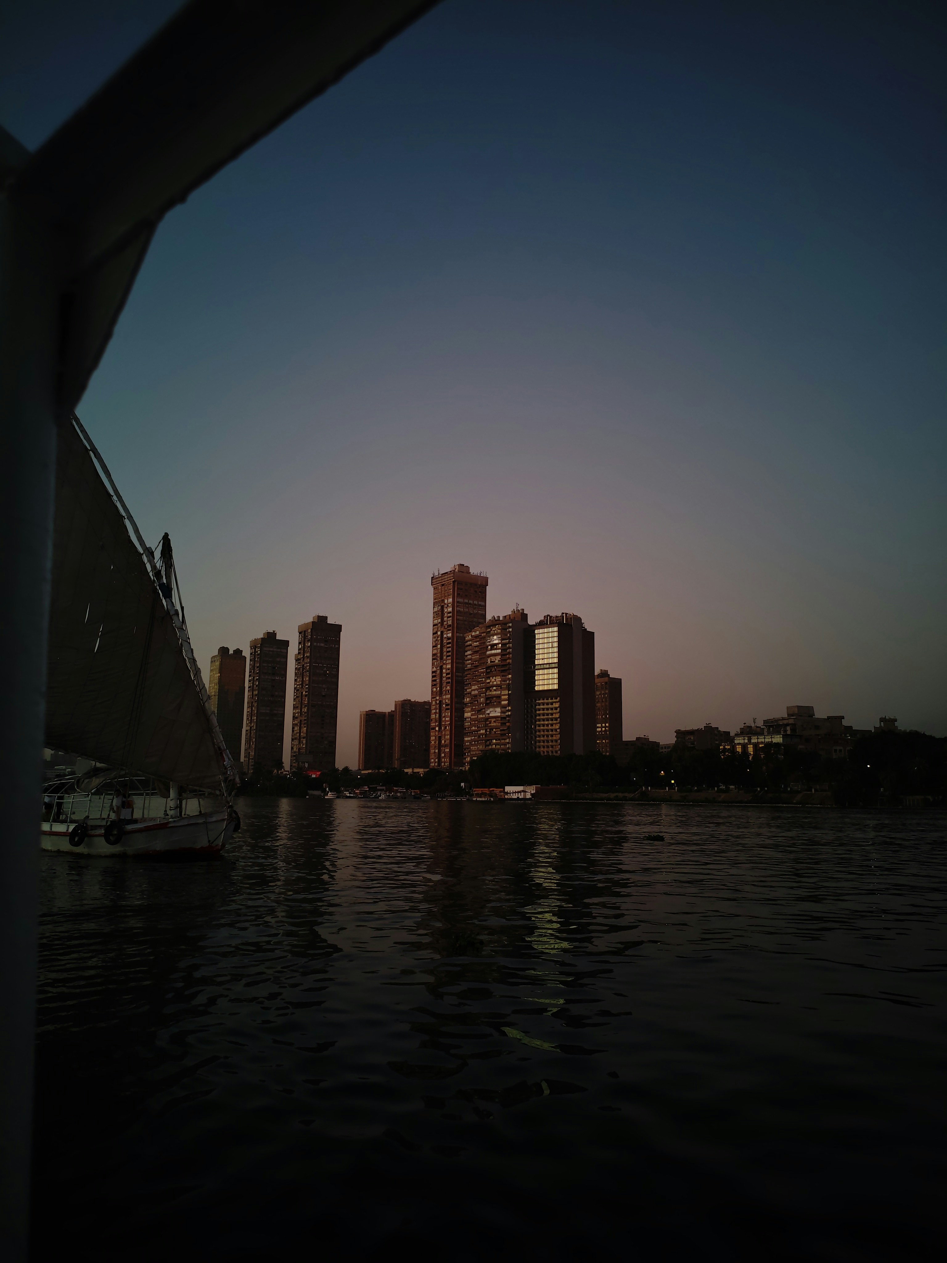 Silhouetted skyscrapers along a riverbank at twilight, with a sailboat partially visible in the foreground.
