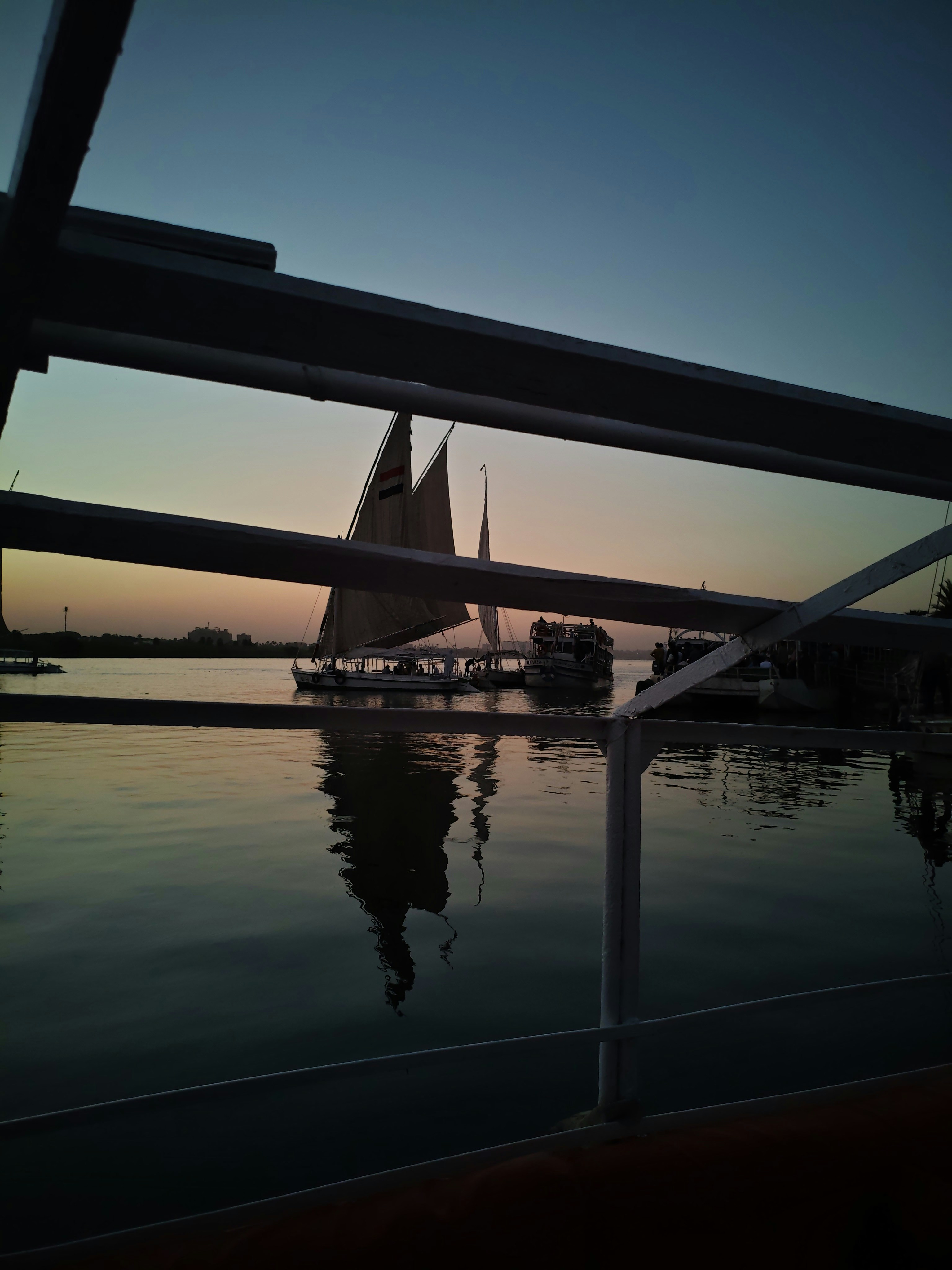 Traditional sailboats navigating a tranquil waterway at twilight, framed by a boat's railing. The calm waters reflect the fading light.