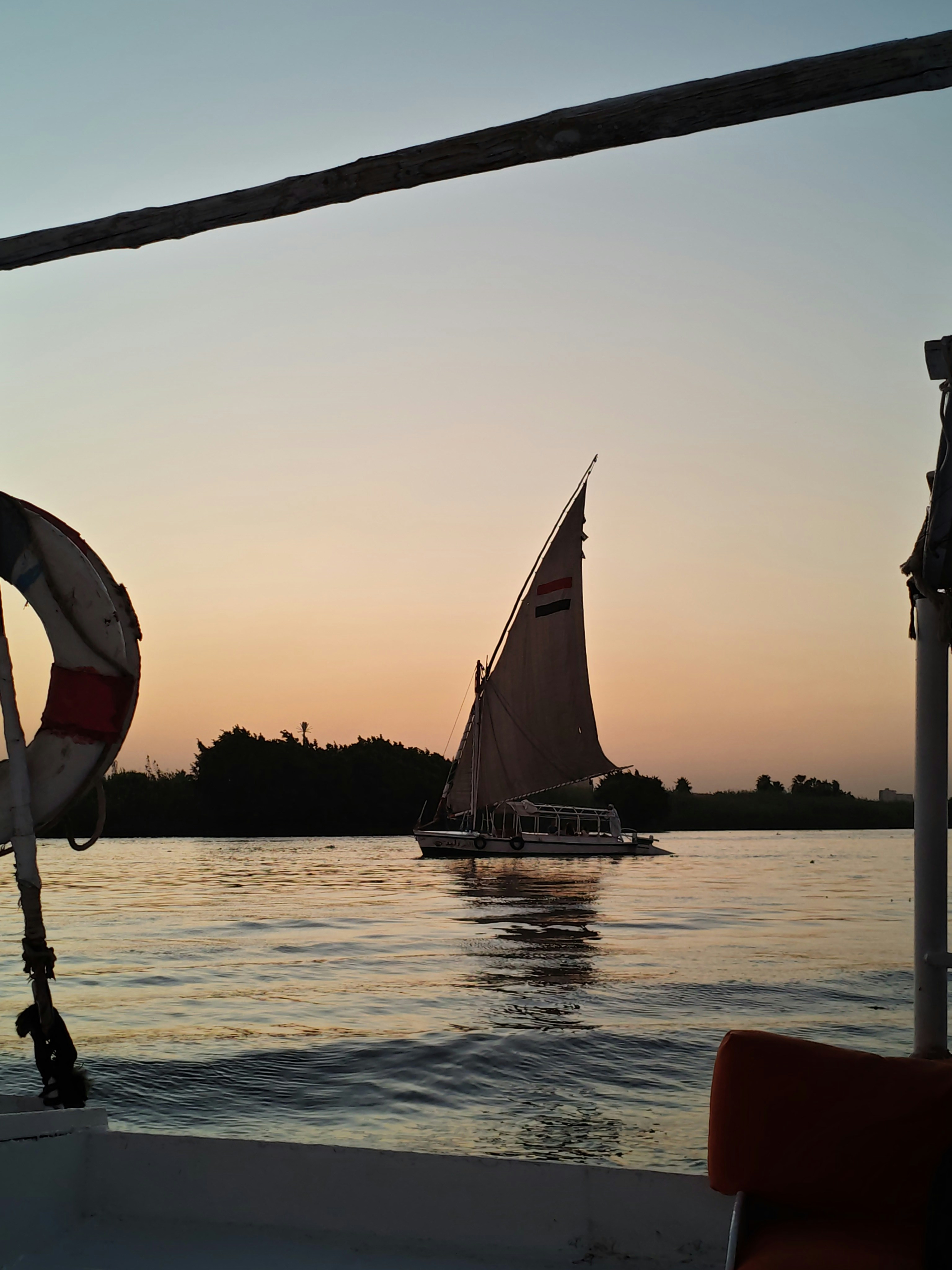 A traditional sailboat glides across a serene waterway at sunset, framed by a lifebuoy and silhouetted trees. The soft hues of dusk create a tranquil atmosphere.