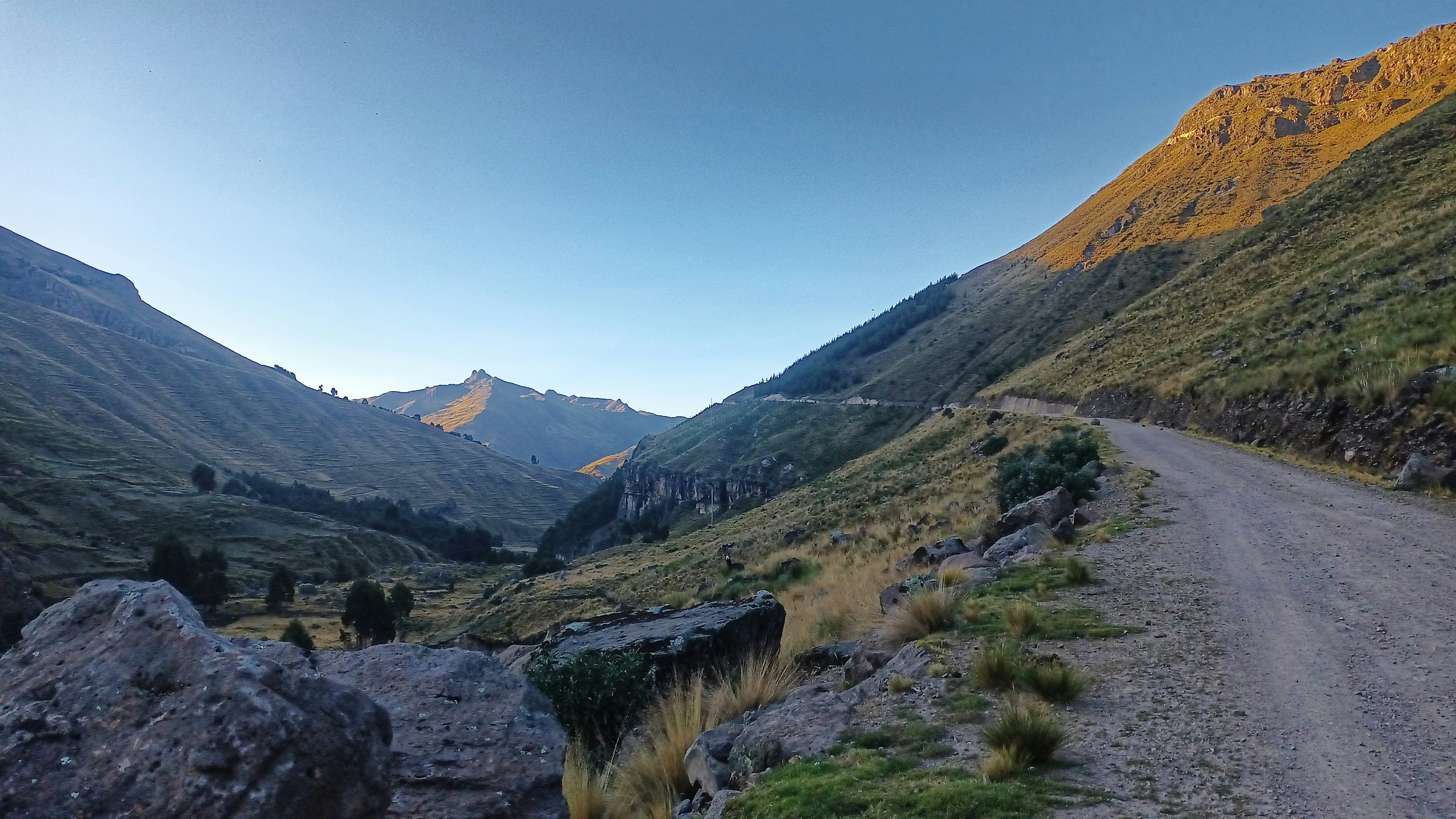 A winding dirt path meanders through lush green hills and rocky formations, leading to distant mountains under a clear blue sky.