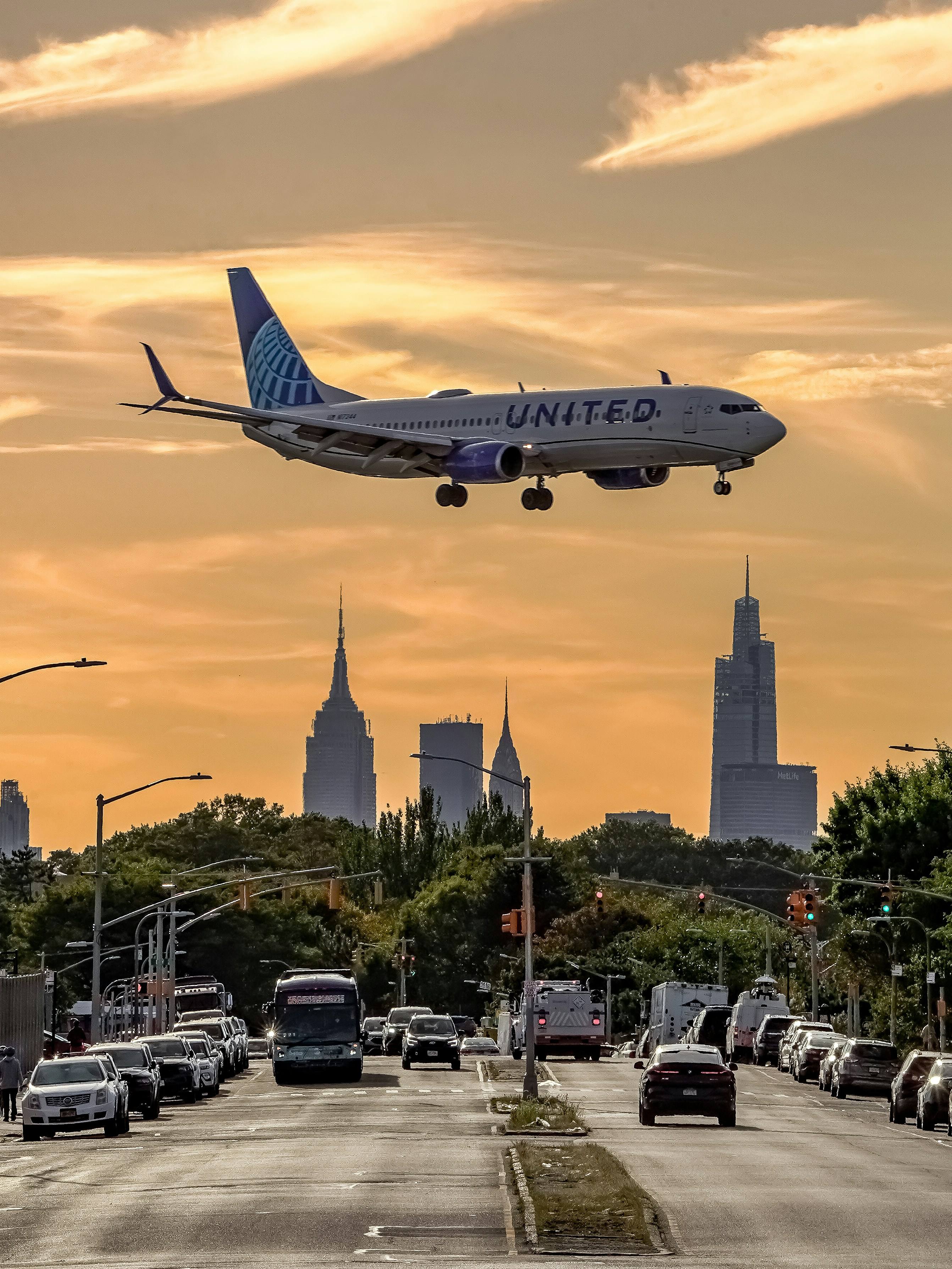 A United Airlines plane descends over a bustling city street at sunset, silhouetted against iconic skyscrapers.