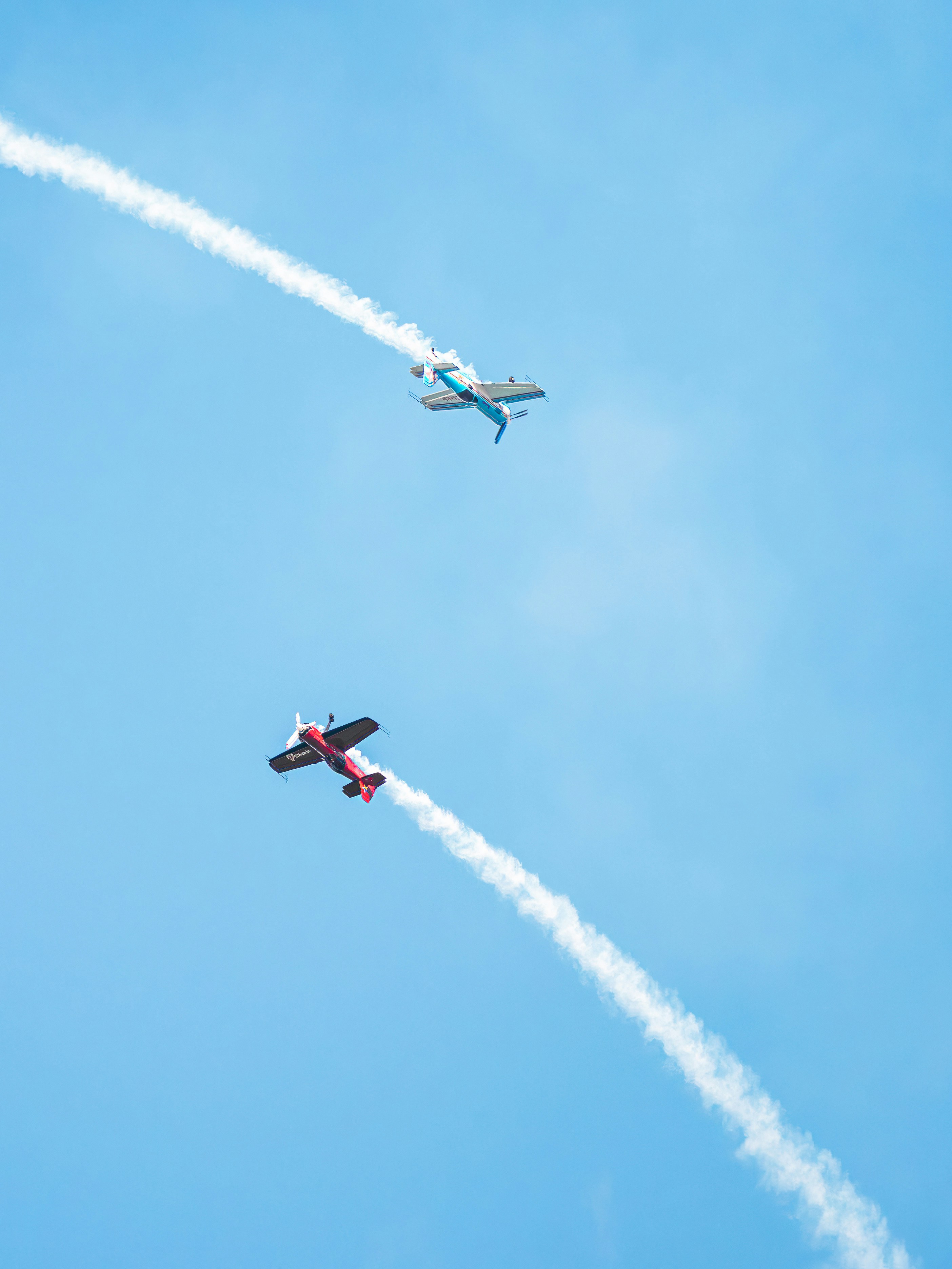 Two acrobatic planes performing synchronized maneuvers, leaving white contrails against a clear blue sky.