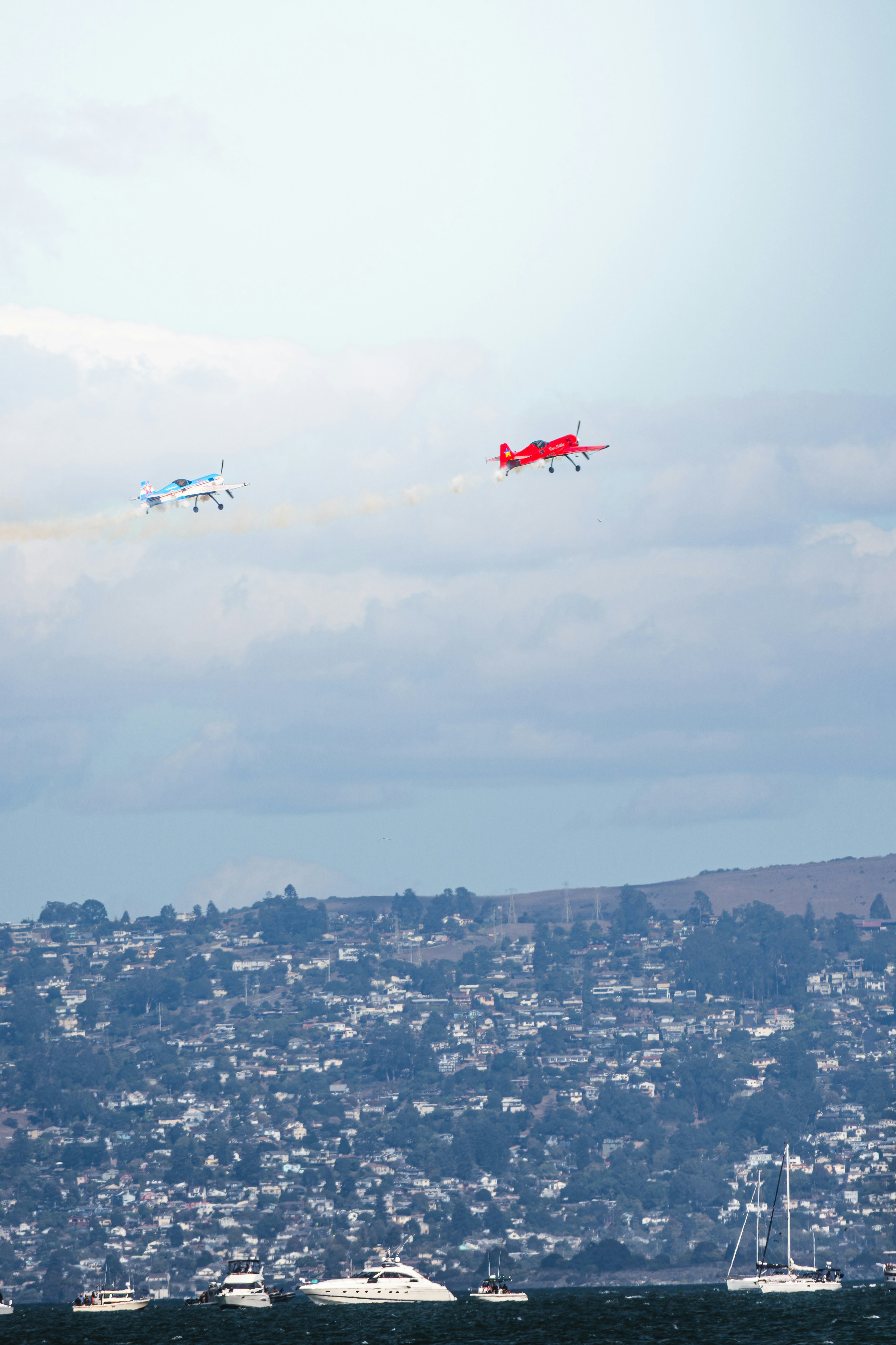 Two aerobatic planes perform synchronized maneuvers in a cloudy sky above a coastal cityscape. The vibrant red and blue aircraft create a dynamic contrast against the backdrop.