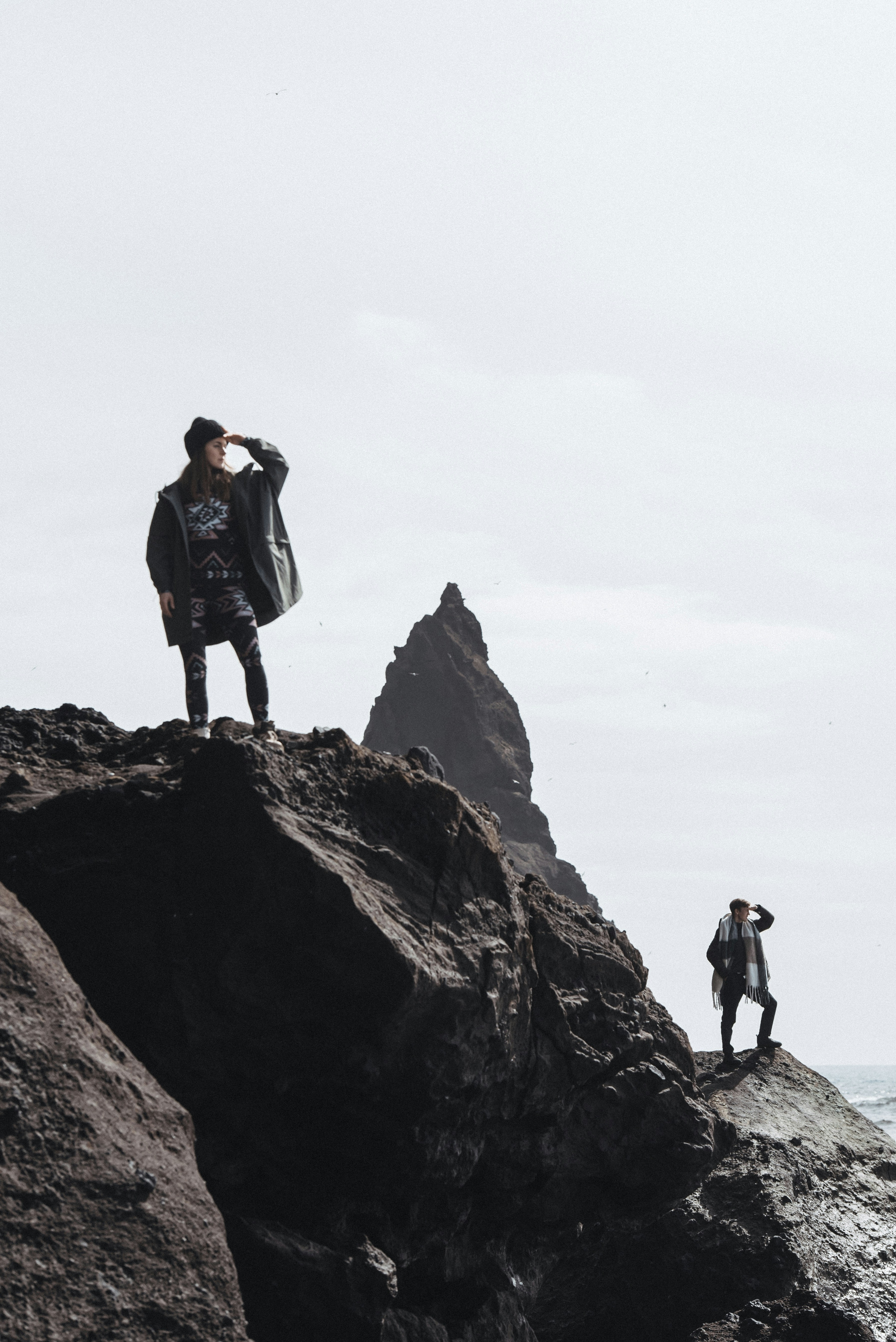 Two people stand on rocky cliffs overlooking the ocean.