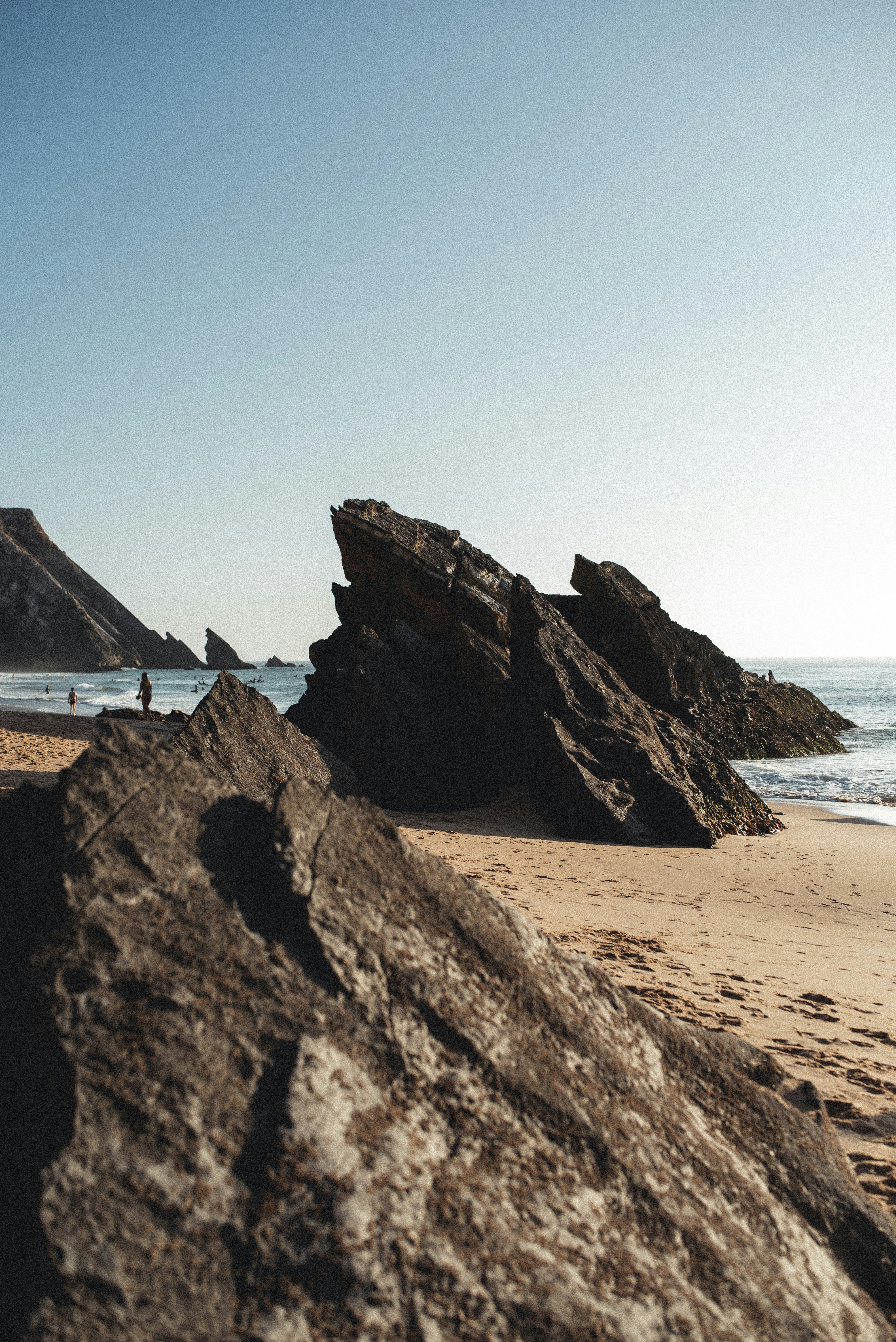 Jagged rock formations rise dramatically from the sandy beach, with gentle waves lapping at their base. A few silhouettes of beachgoers can be seen in the distance.