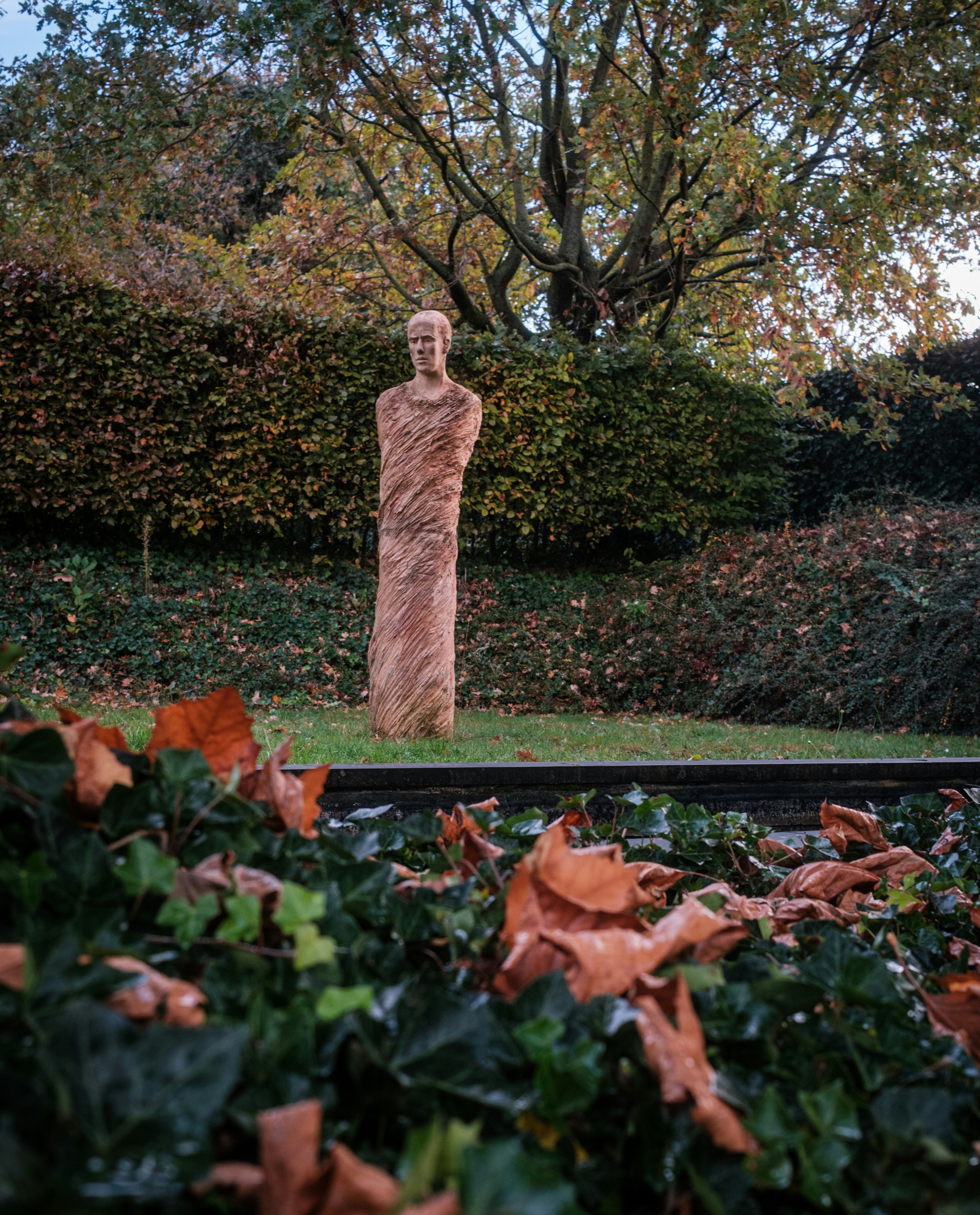 Wooden sculpture of a figure standing amidst fallen leaves and greenery in a serene garden setting.