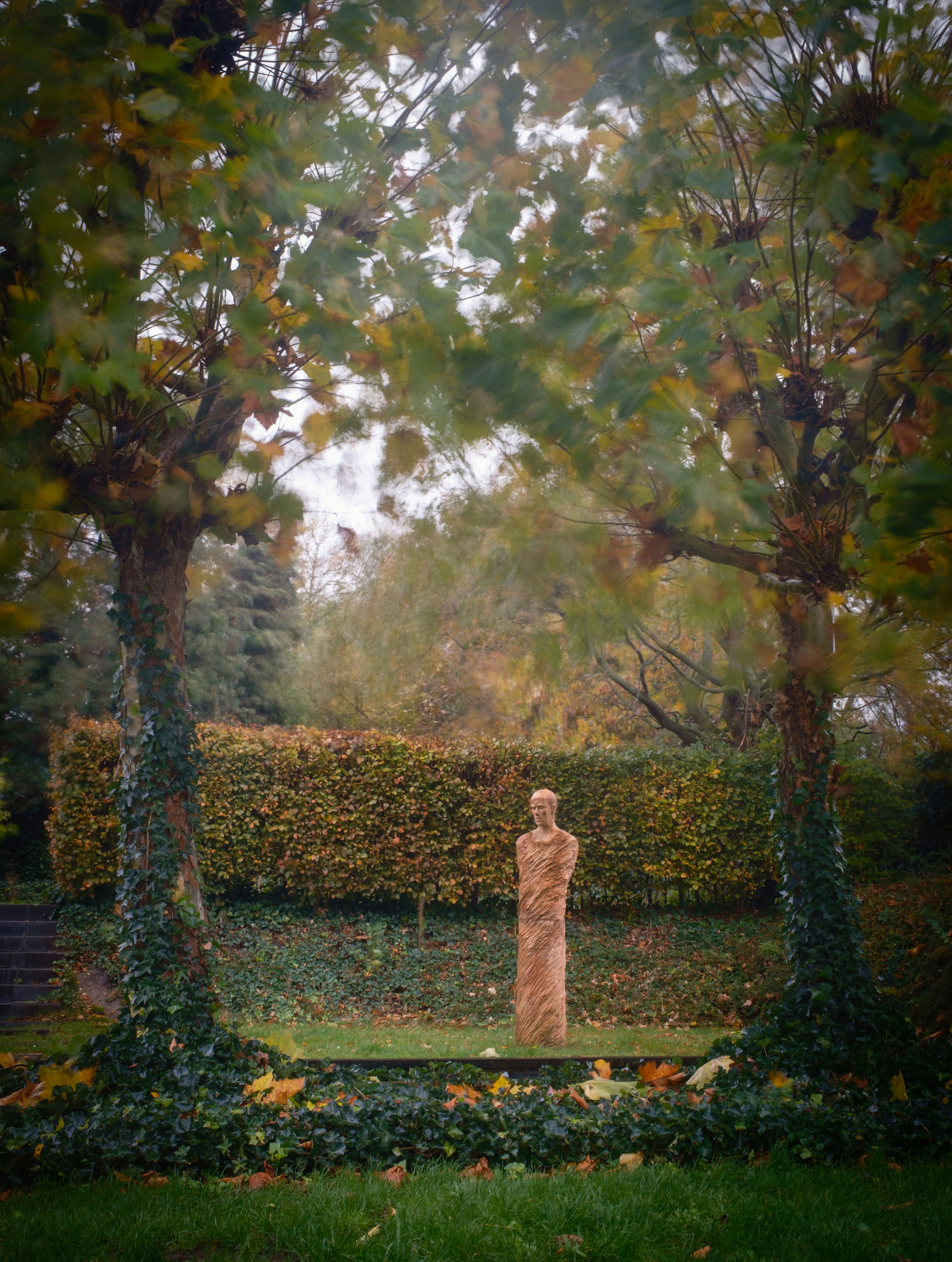A solitary statue stands amidst vibrant autumn foliage, framed by trees and ivy, evoking a sense of tranquility and reflection.