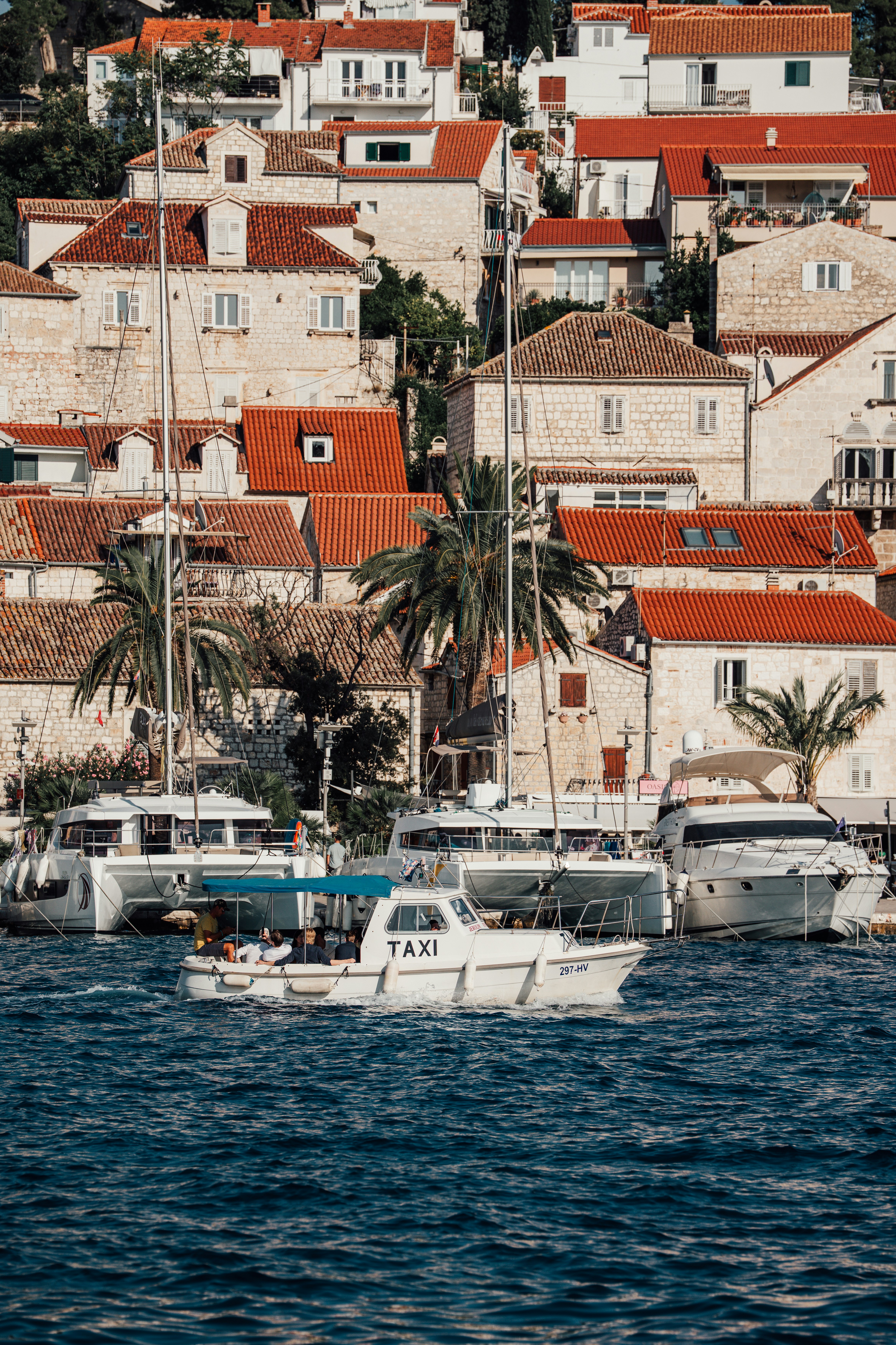 White taxi boat navigating the harbor amidst a backdrop of charming stone houses with red roofs and palm trees. 