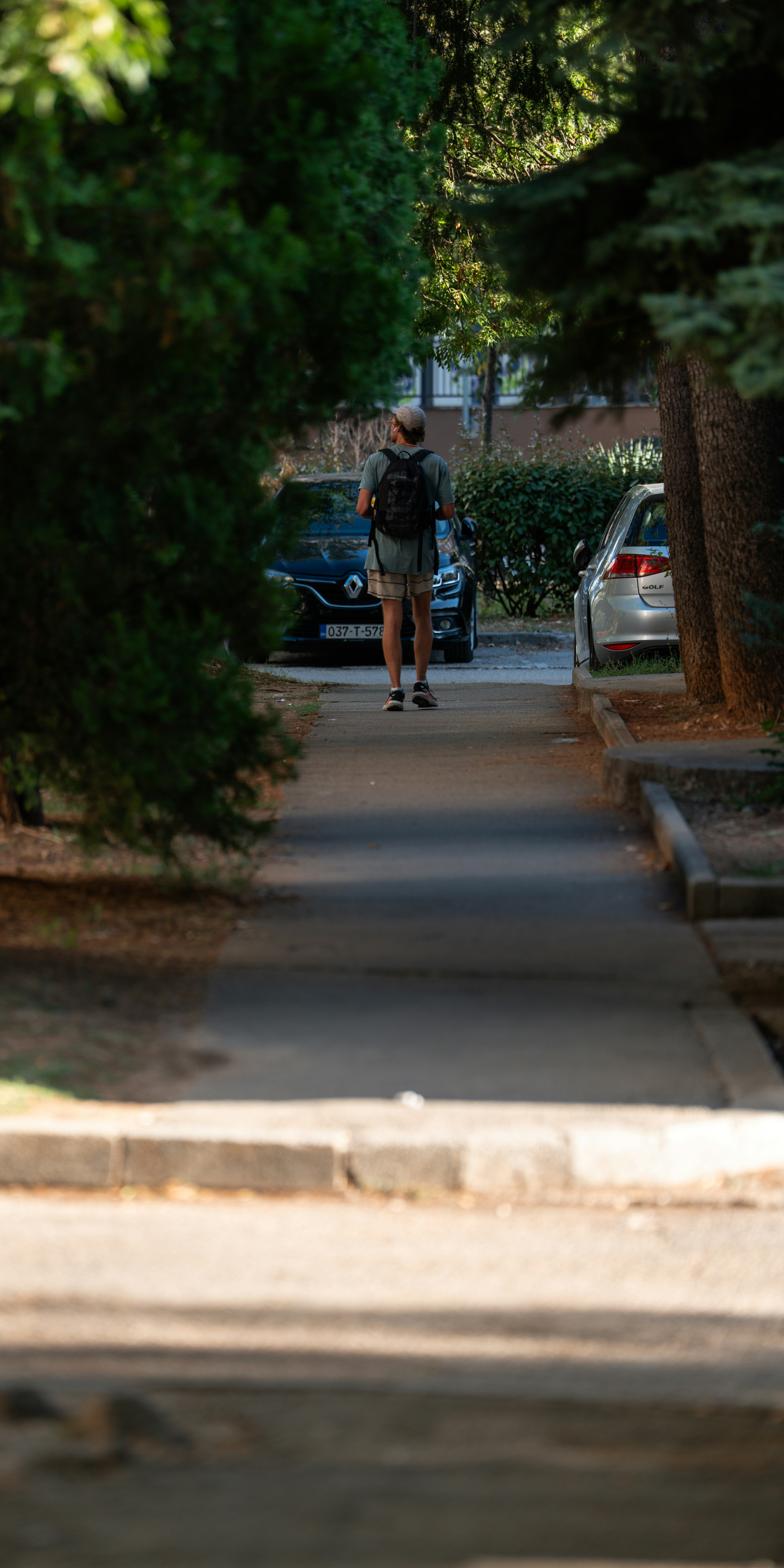 Individual walking along a tree-lined path, framed by lush greenery and parked cars in the background.