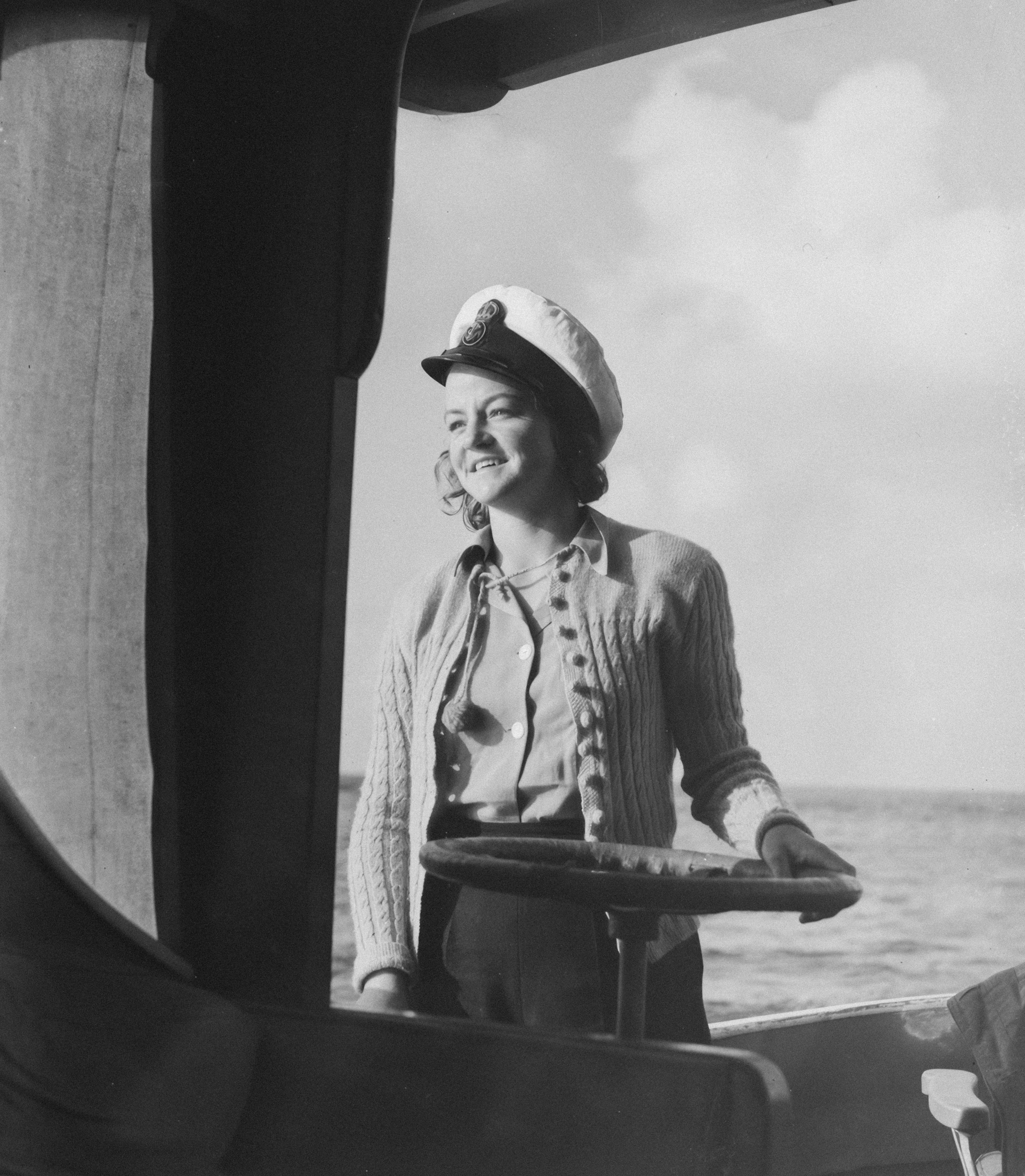 This photograph was taken by Tudor Washington Collins (b.1898, d.1970) in possibly in 1946. This scene depicts a portrait (photographed from inside) of a woman wearing a Naval cap and holding the steering wheel of a boat, with the sea showing in the background. Credit: Shared by Auckland War Memorial Museum, Tāmaki Paenga Hira, as part of the Tudor Collins collection. Rights: No known copyright restrictions. Reference: PH-2013-7-TC-B504-16. For more details, please visit: www.aucklandmuseum.com/discover/collections/record/1054812 | Woman wearing a captain's hat at the helm
