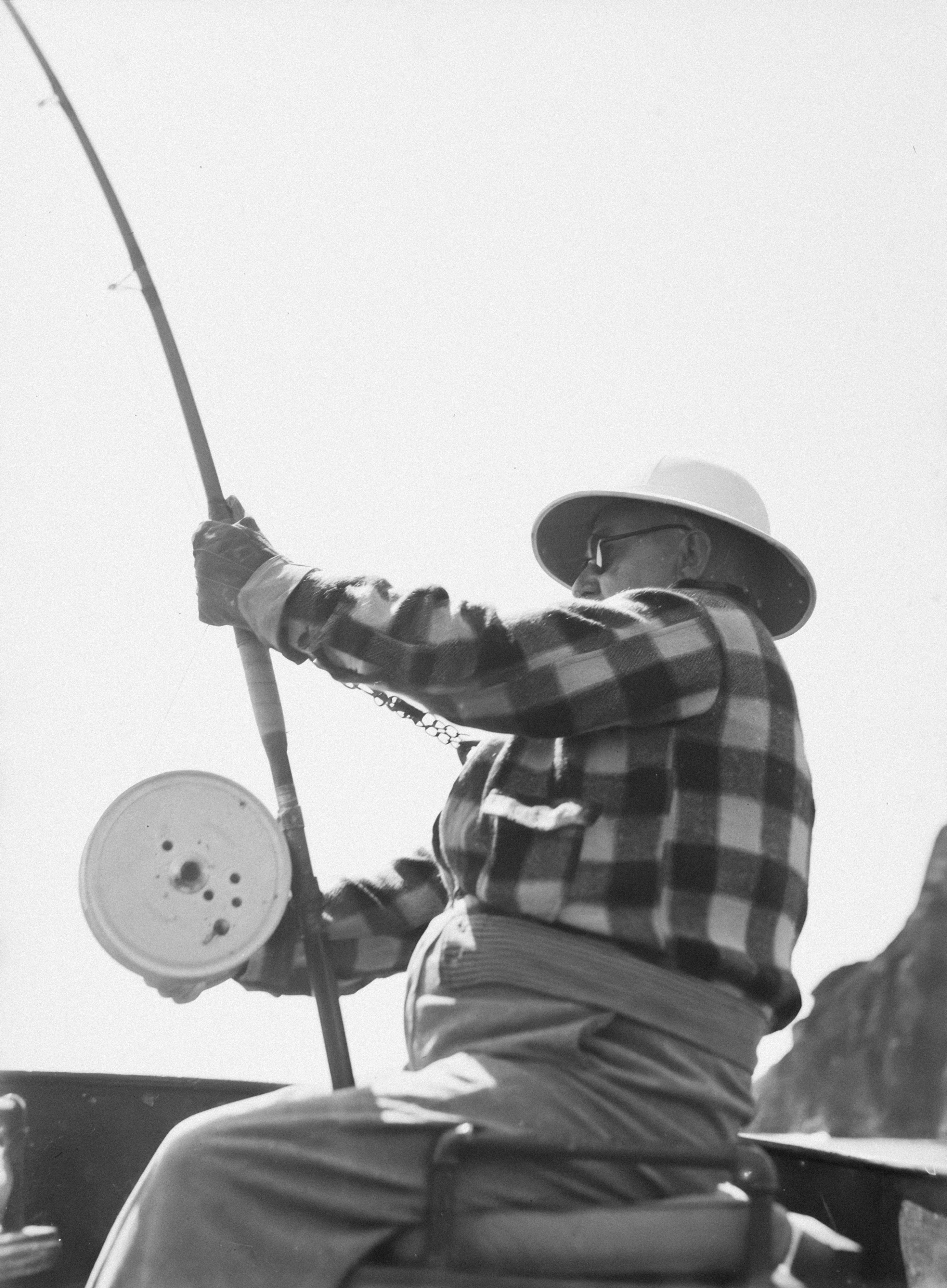Elderly fisherman skillfully casting his line from a boat, wearing a plaid shirt and wide-brimmed hat. The scene captures the essence of a tranquil fishing experience.