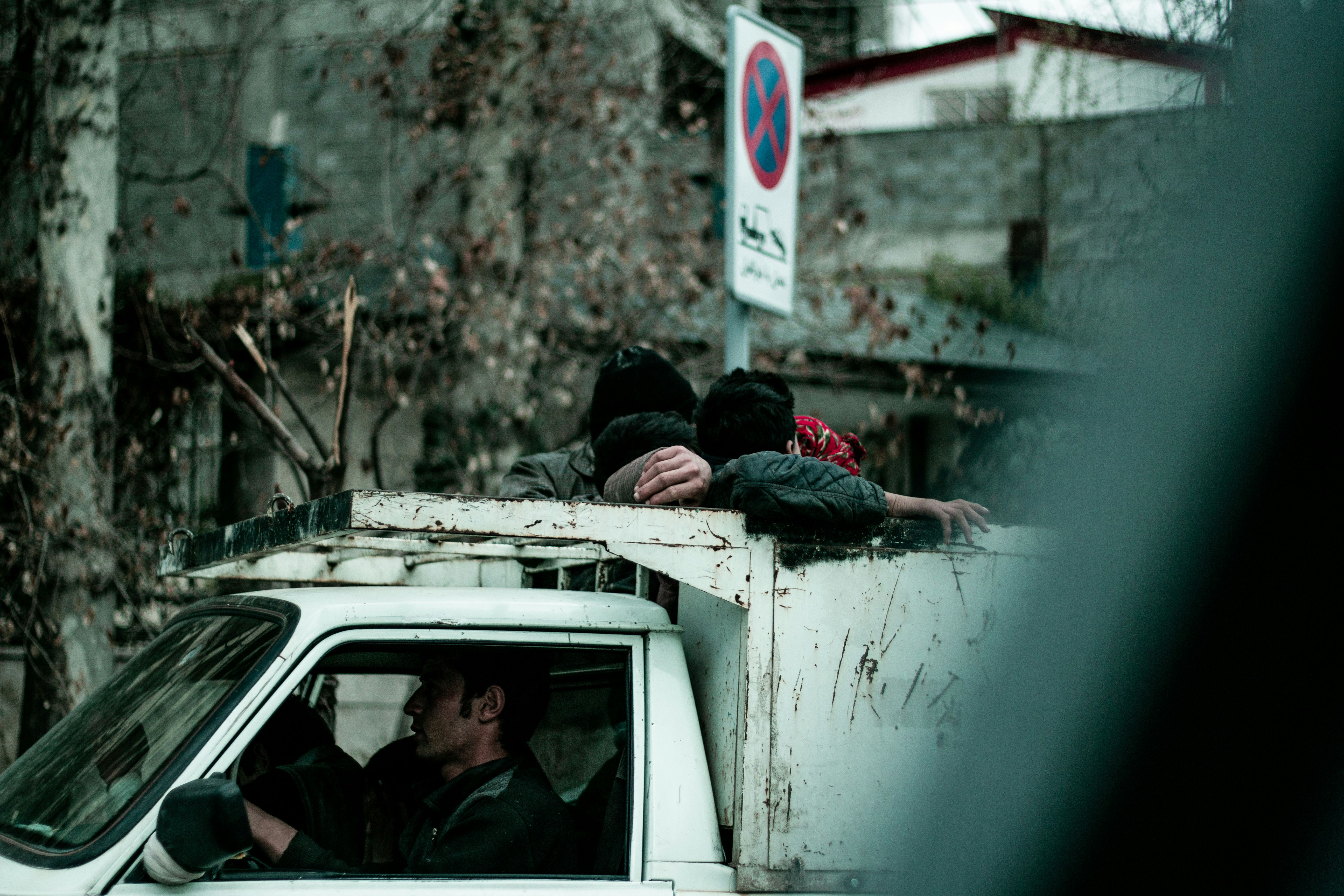 Two figures share a moment of intimacy atop a truck, while a driver remains focused on the road. A no-parking sign looms in the background.