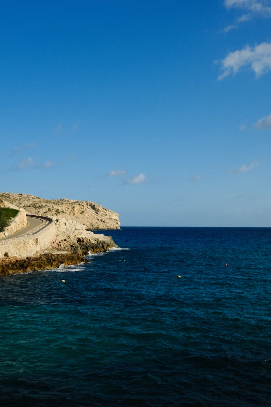 A winding road hugs pale rocky cliffs beside deep blue water under a clear sky with a few small clouds.