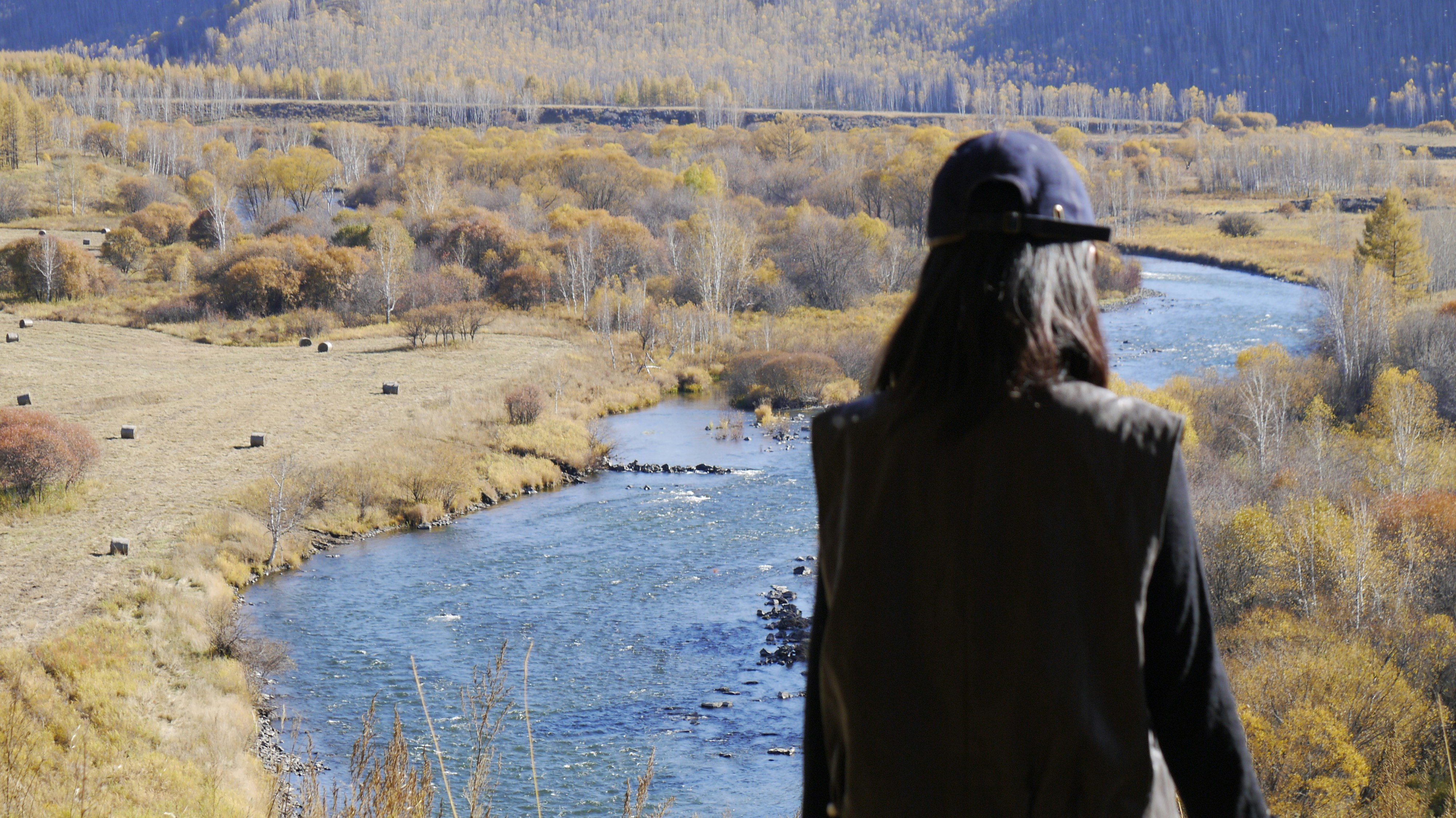 A person gazes over a winding river surrounded by golden autumn foliage and distant hills. The scene captures the tranquil beauty of nature in transition.