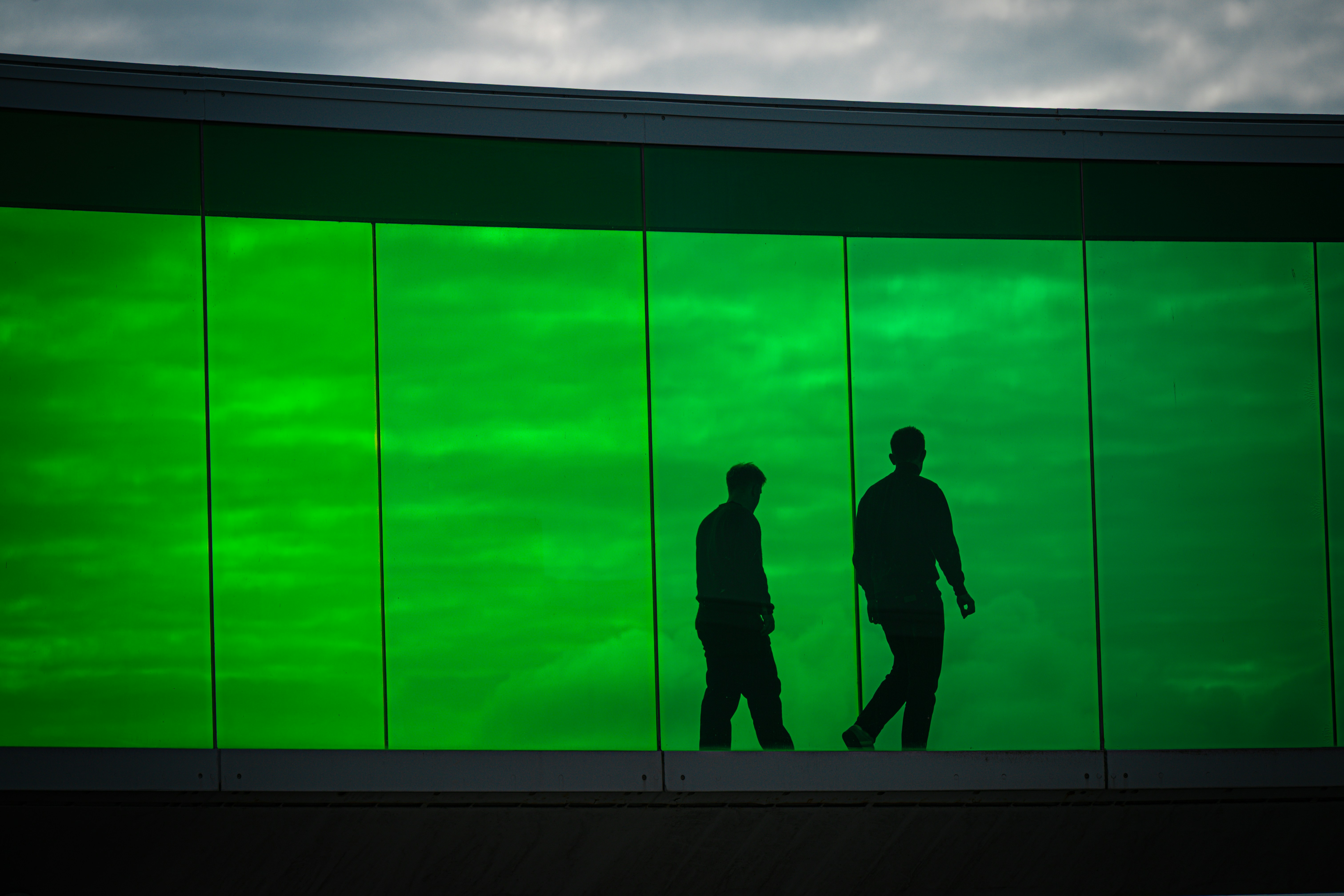 Two Silhouettes Trek Across a Lime-Green Glass Corridor