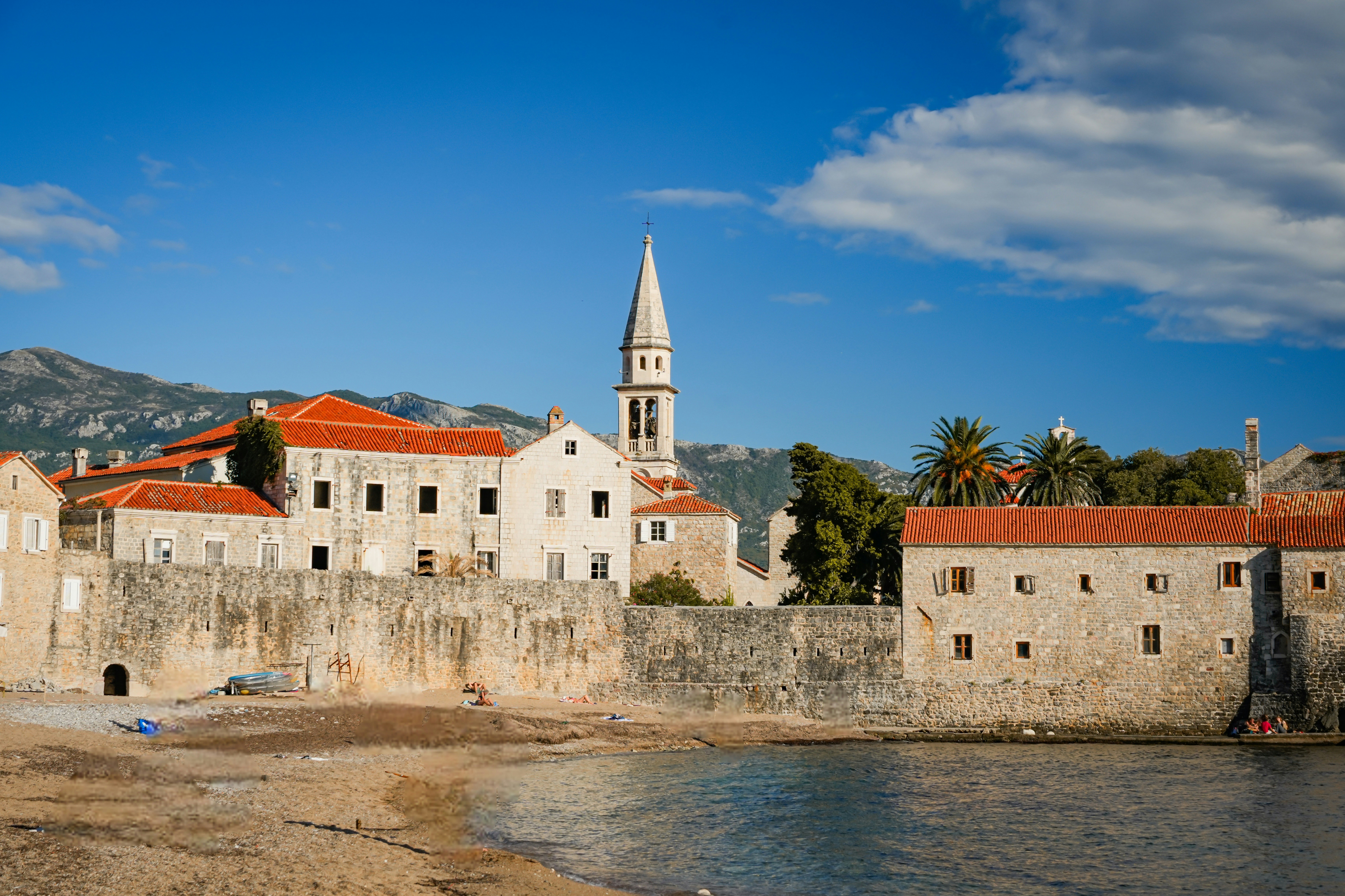 Historic buildings with red roofs and a tall steeple overlook a serene beach, framed by mountains and palm trees.