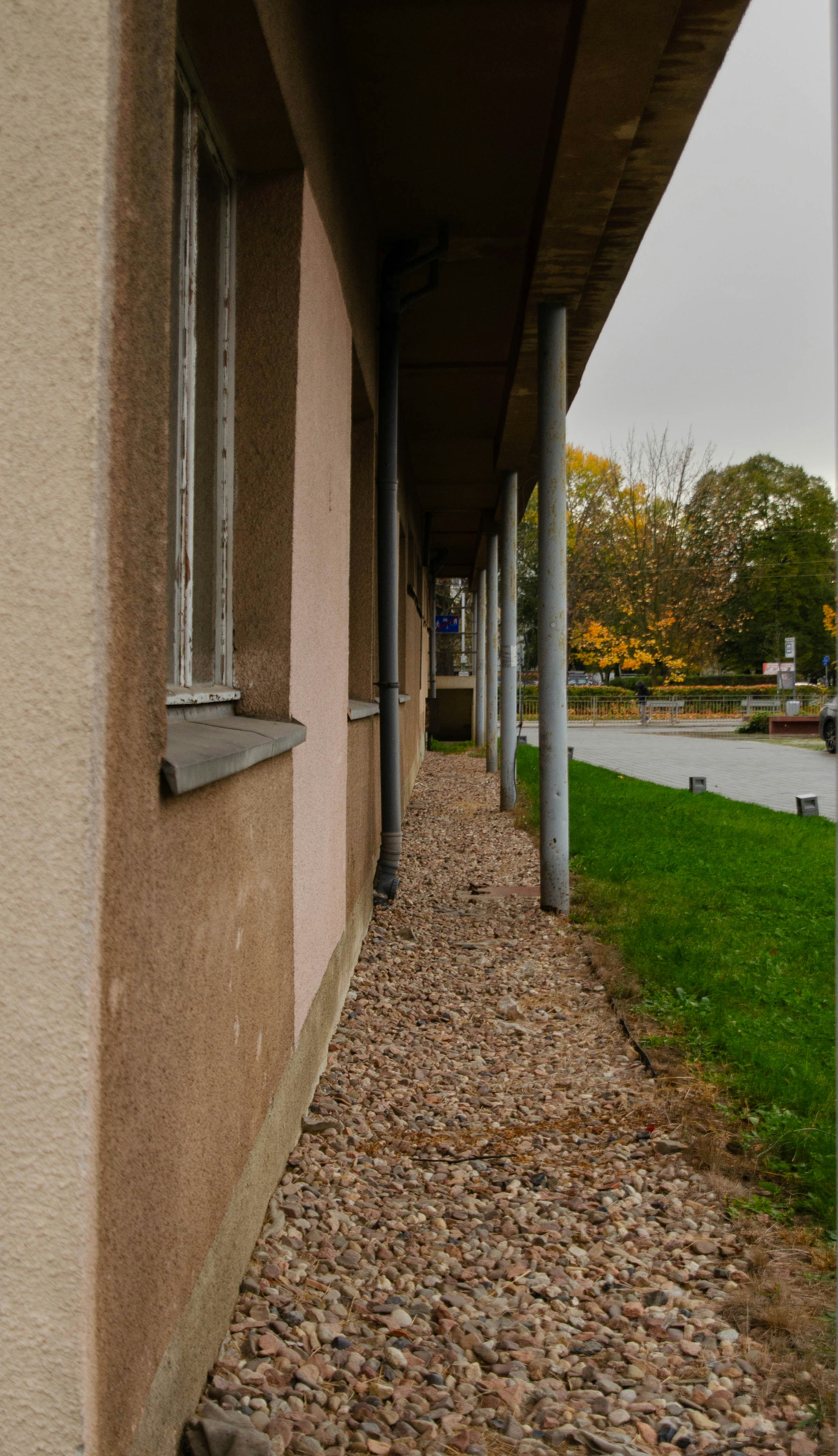 Narrow gravel pathway alongside a building, framed by muted colors and distant autumn foliage. The scene evokes a sense of solitude.
