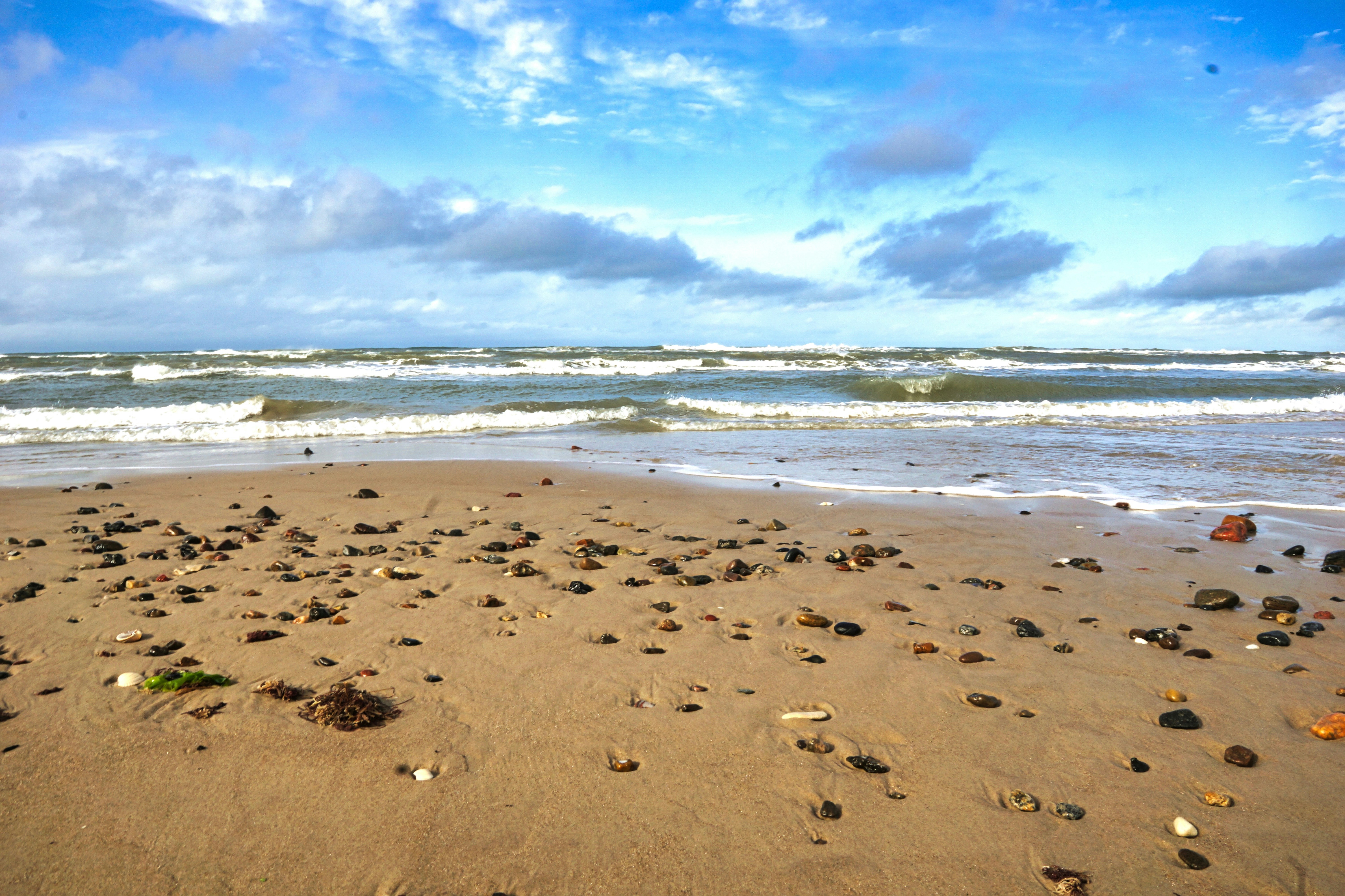 A sandy beach dotted with various pebbles and shells, where gentle waves lap at the shore under a partly cloudy sky.