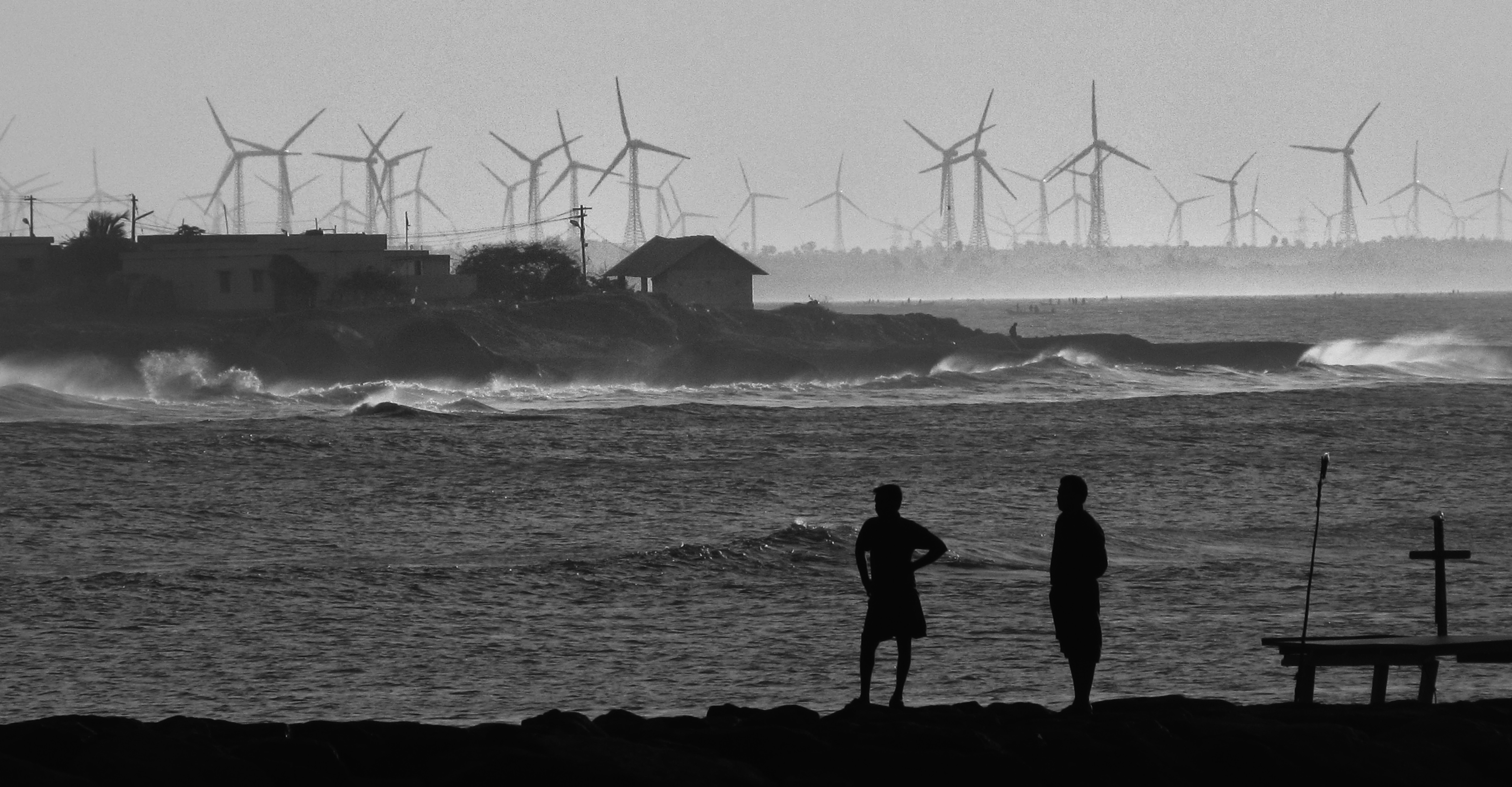 Two silhouetted figures stand on the shoreline, gazing out at the waves and wind turbines in the distance. The scene captures a moment of reflection amidst nature's elements.