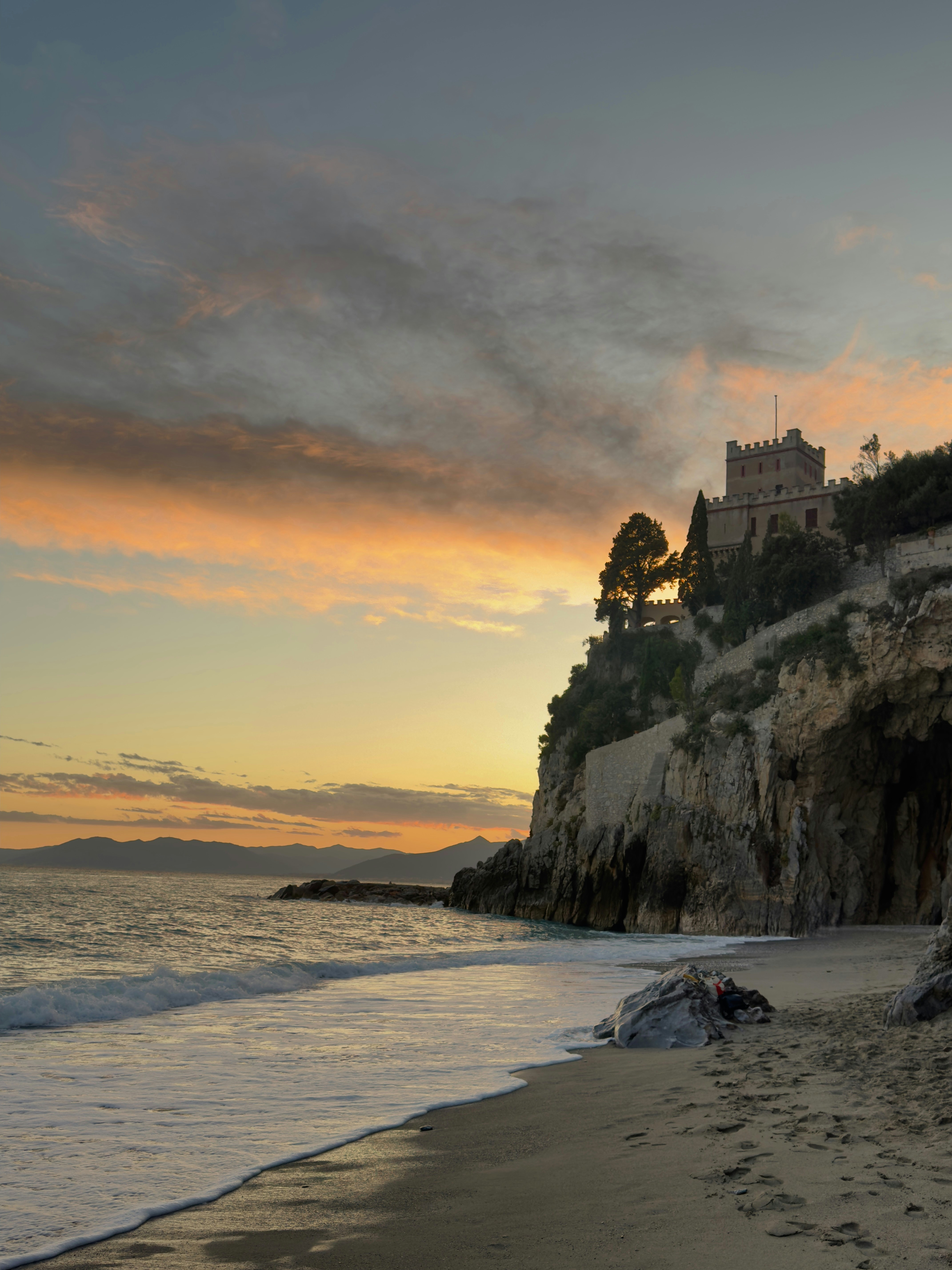 A coastal scene featuring a grand castle perched on a rocky cliff, illuminated by the warm hues of a setting sun, with gentle waves lapping at the sandy beach.