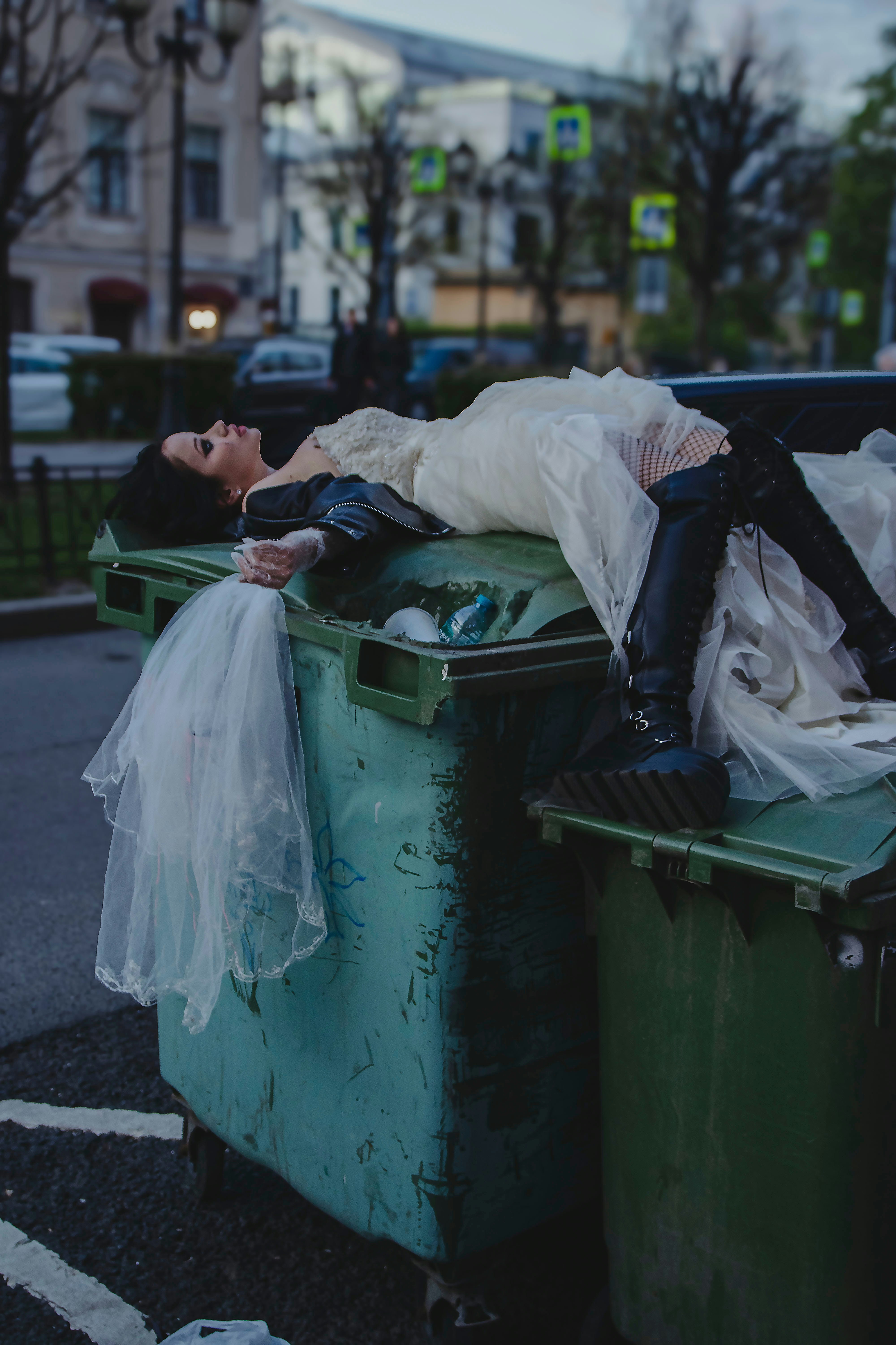 A figure draped in a wedding dress lies across two green dumpsters, juxtaposing beauty and decay in an urban setting.