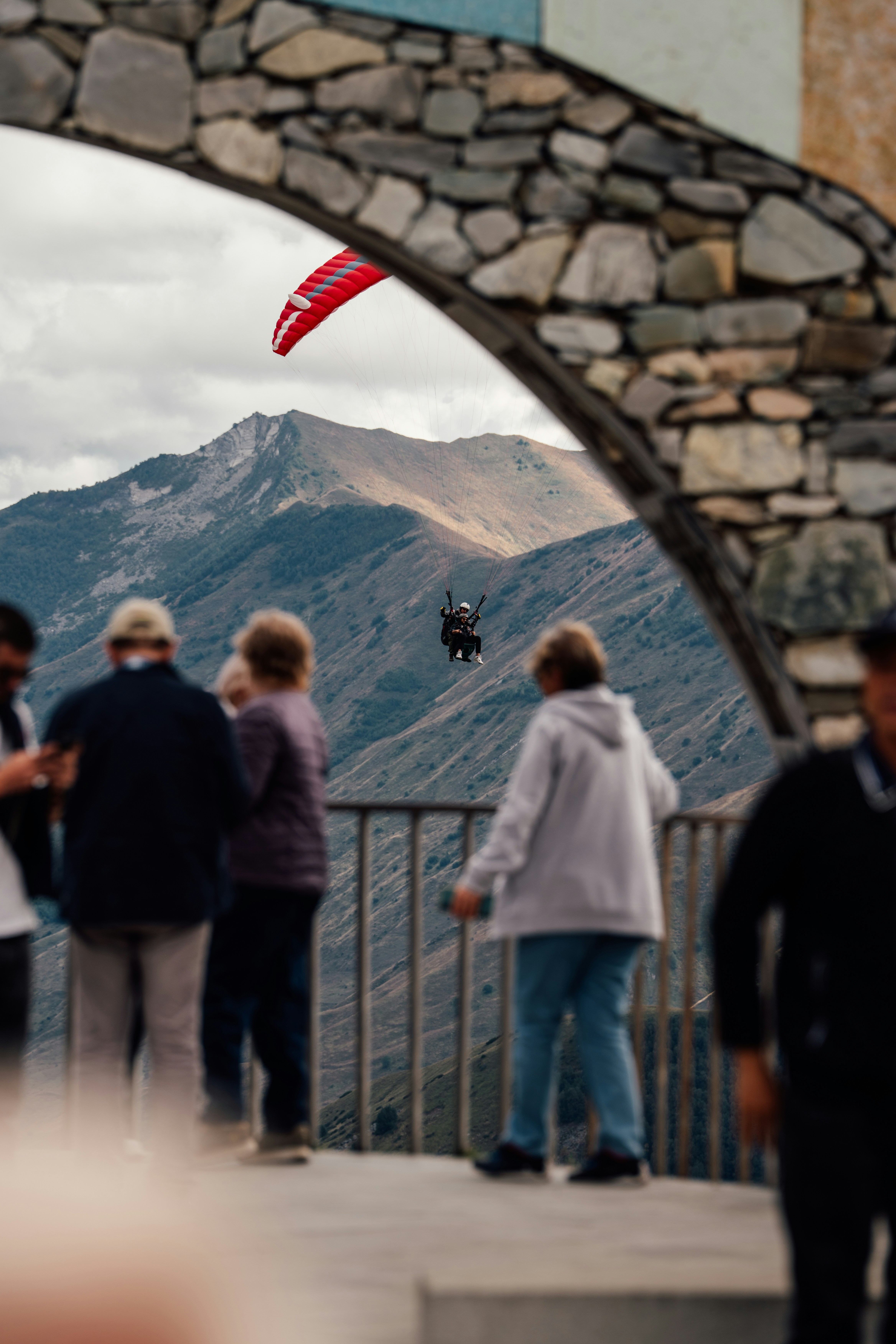 A paraglider gracefully descends through a stone archway, surrounded by onlookers on a scenic overlook. The backdrop features majestic mountains under a cloudy sky.