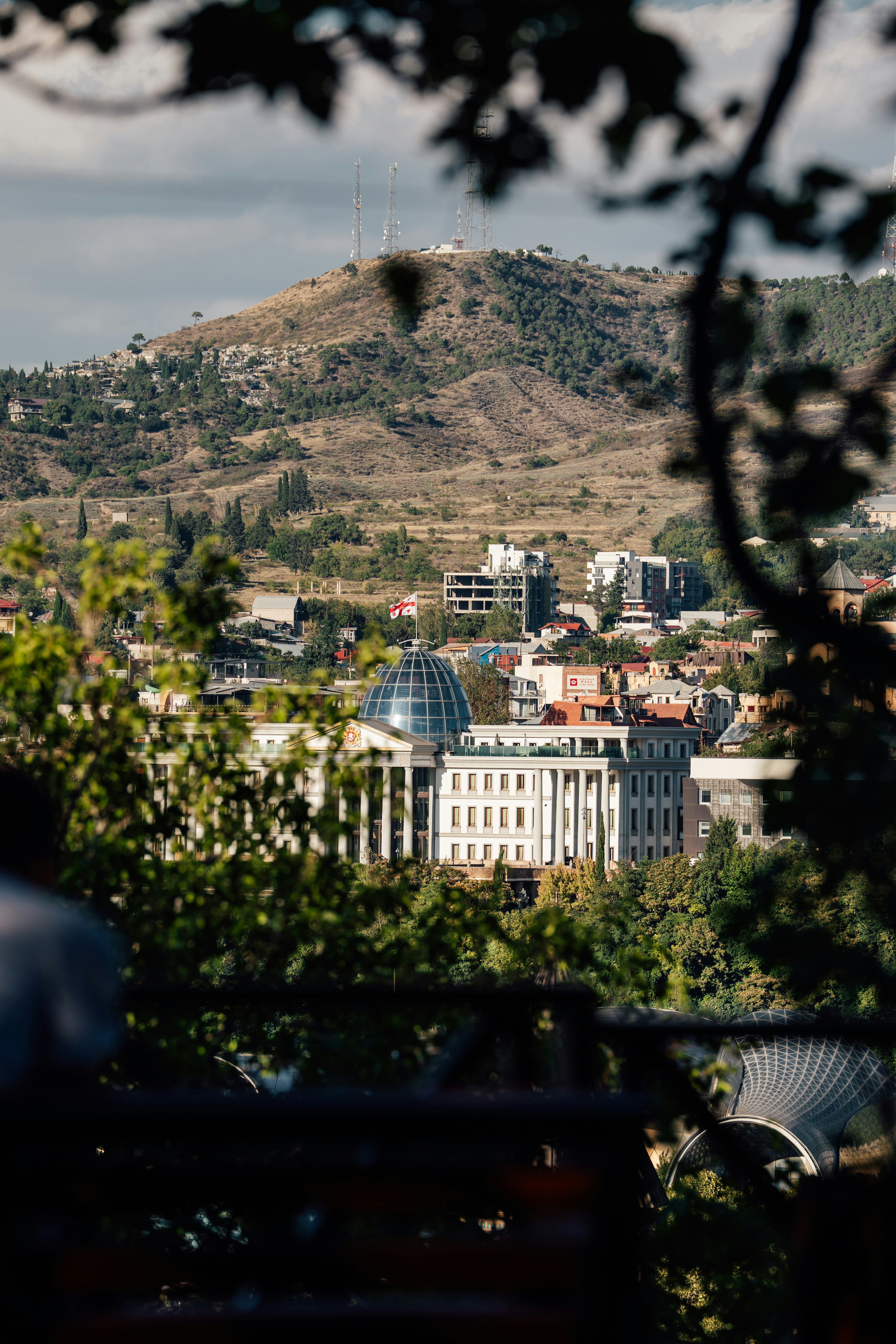 View of a cityscape framed by lush greenery, showcasing a prominent building with a dome against a backdrop of hills. 