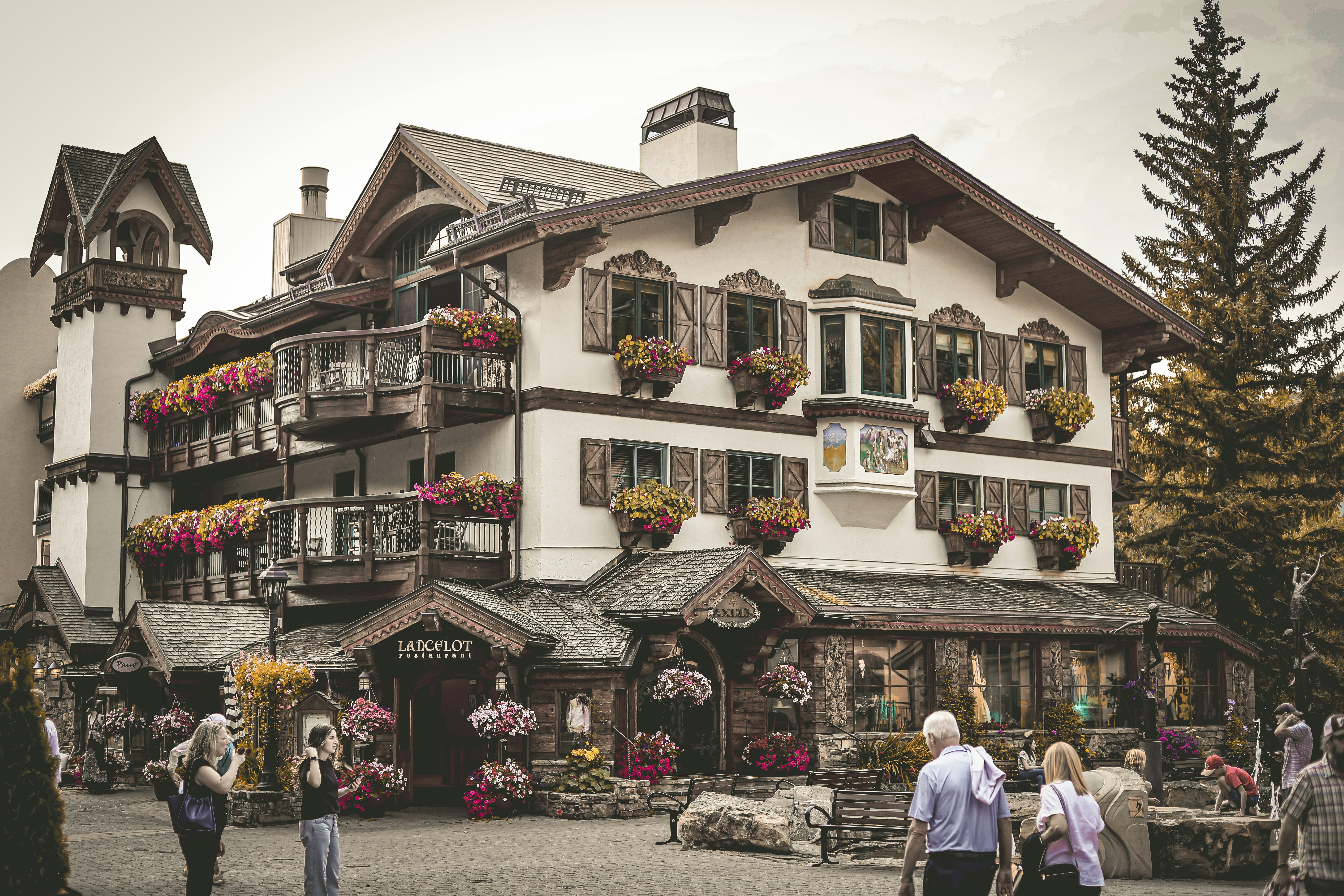 A picturesque alpine lodge adorned with vibrant flower boxes, set against a backdrop of tall trees and a cloudy sky.